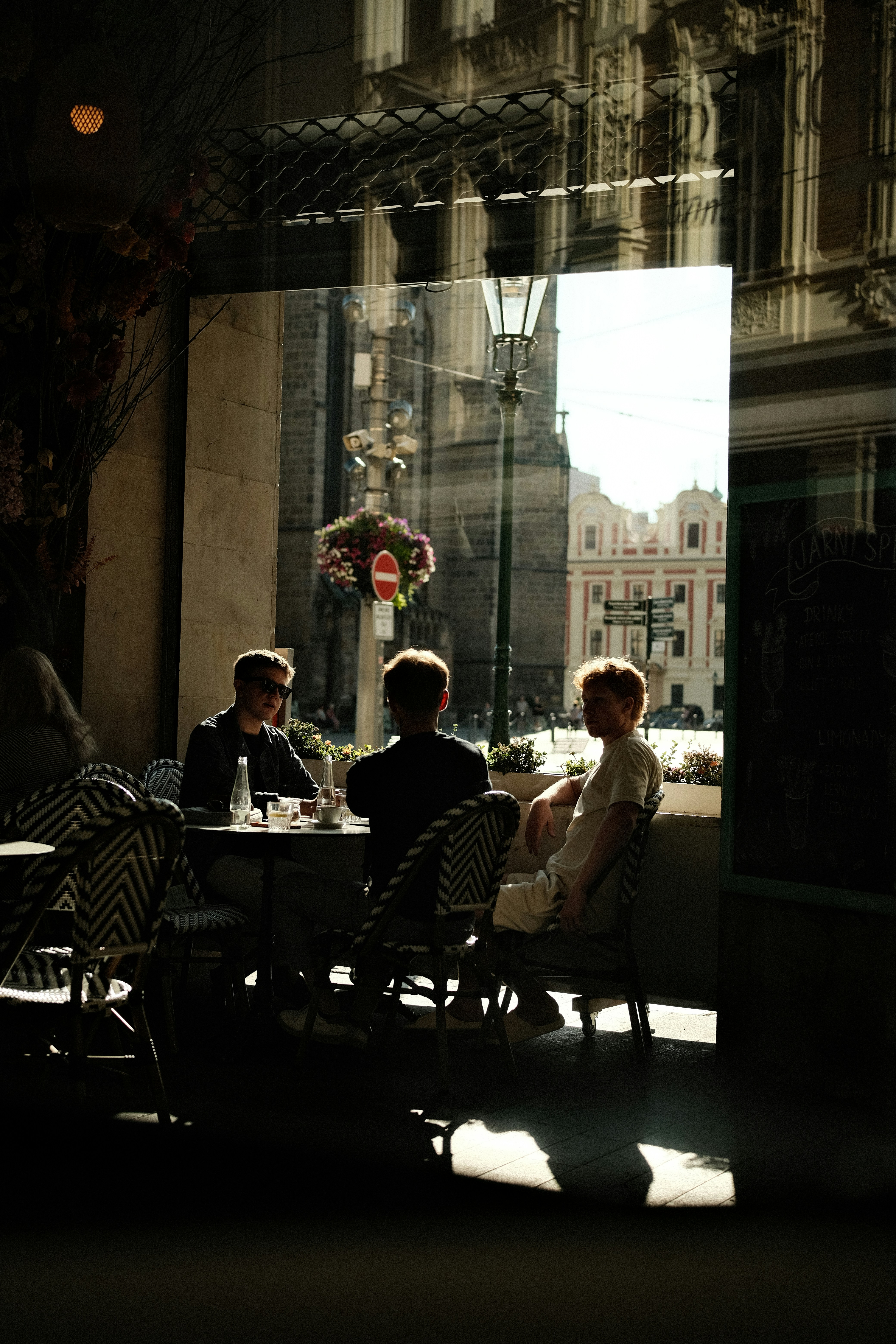 Three individuals sit at a cafe table, engaged in conversation. Sunlight illuminates the outdoor view of a city scene and buildings beyond the glass. | Three people sit at a table outside a cafe.