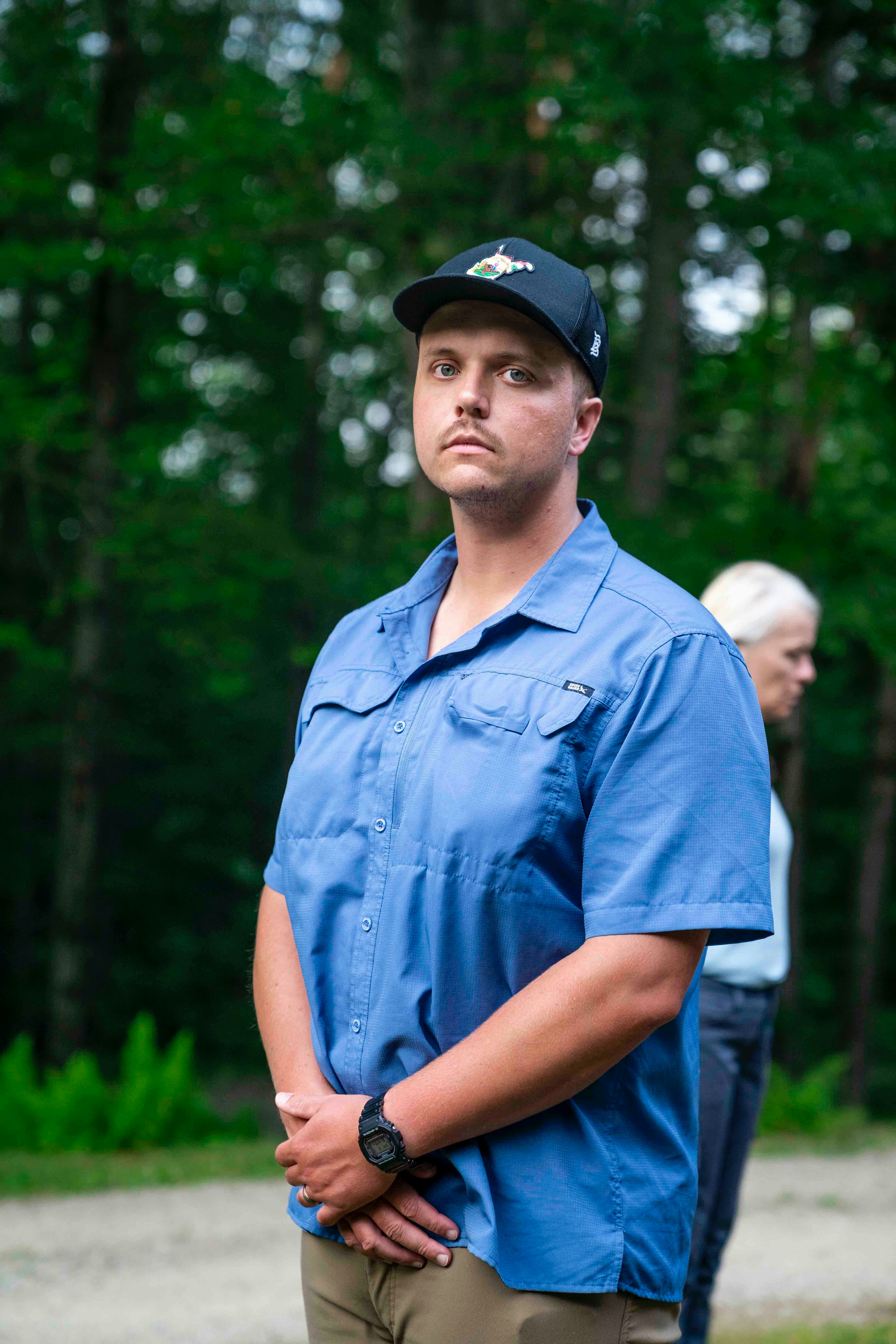 Man in a blue shirt stands confidently in a forested area, with another figure blurred in the background. The lush greenery surrounds them.