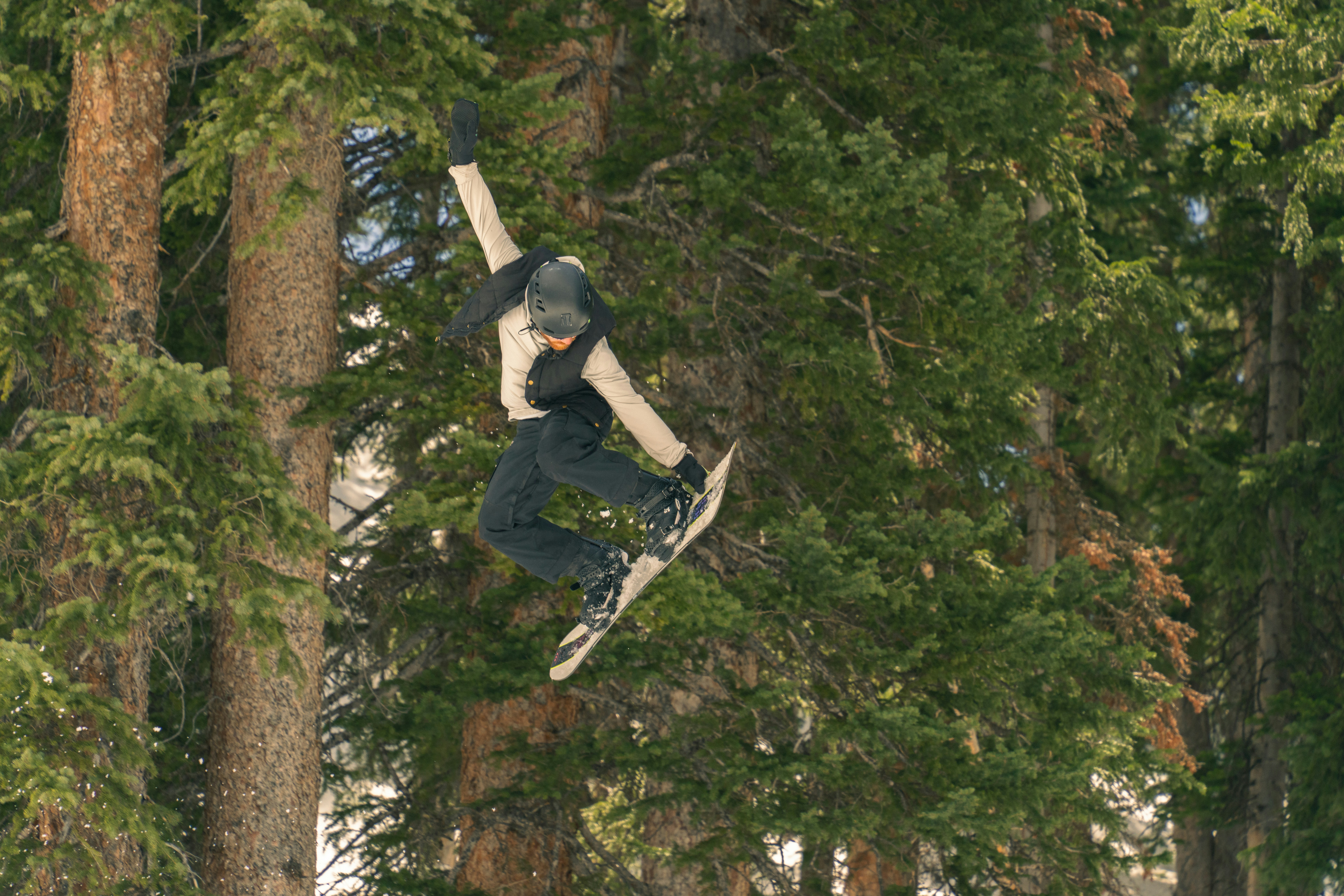 Snowboarder executing a mid-air trick amidst a backdrop of tall evergreen trees.