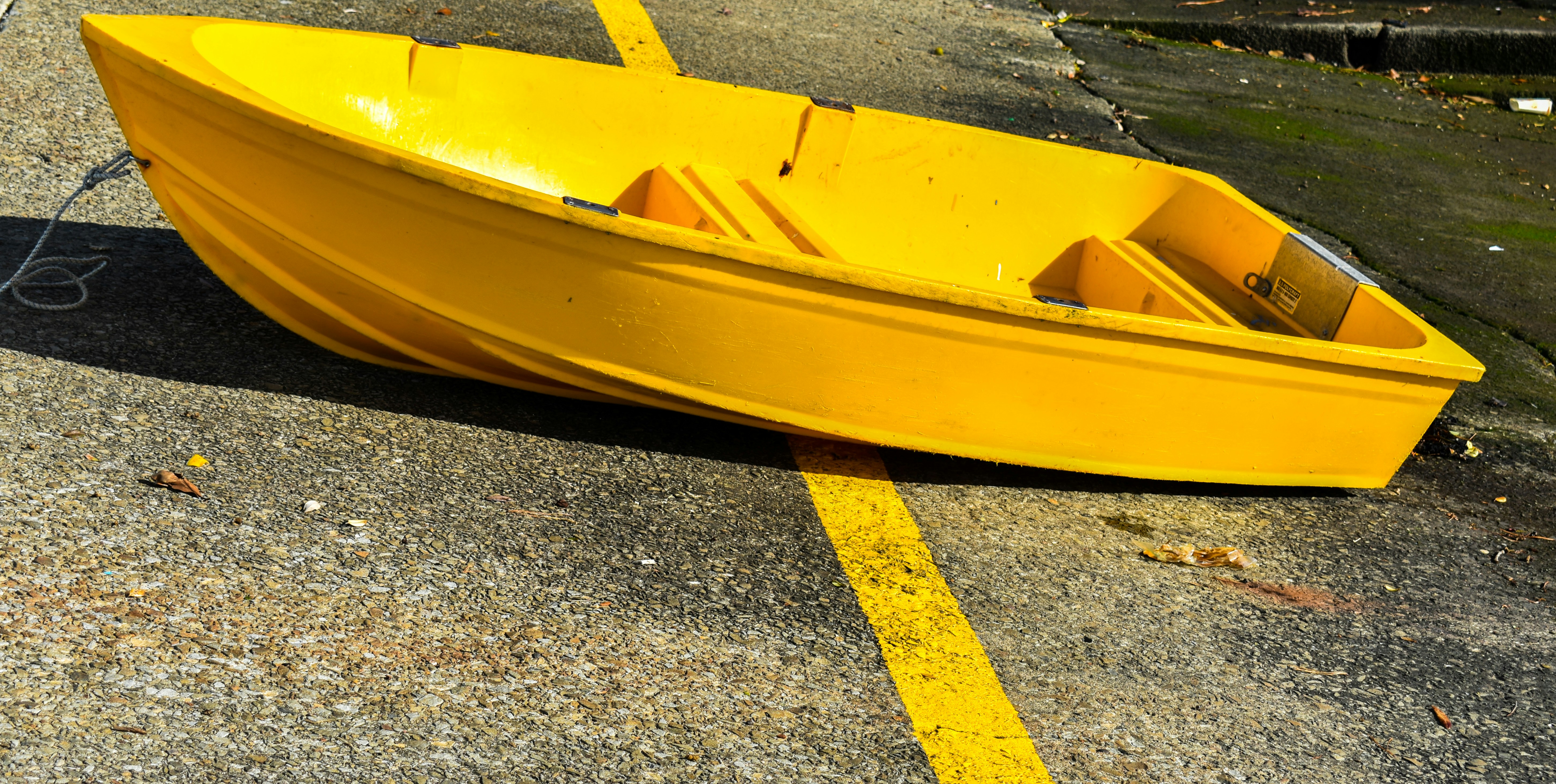A small yellow boat is resting on a yellow line | A bright yellow rowboat rests on a paved surface.