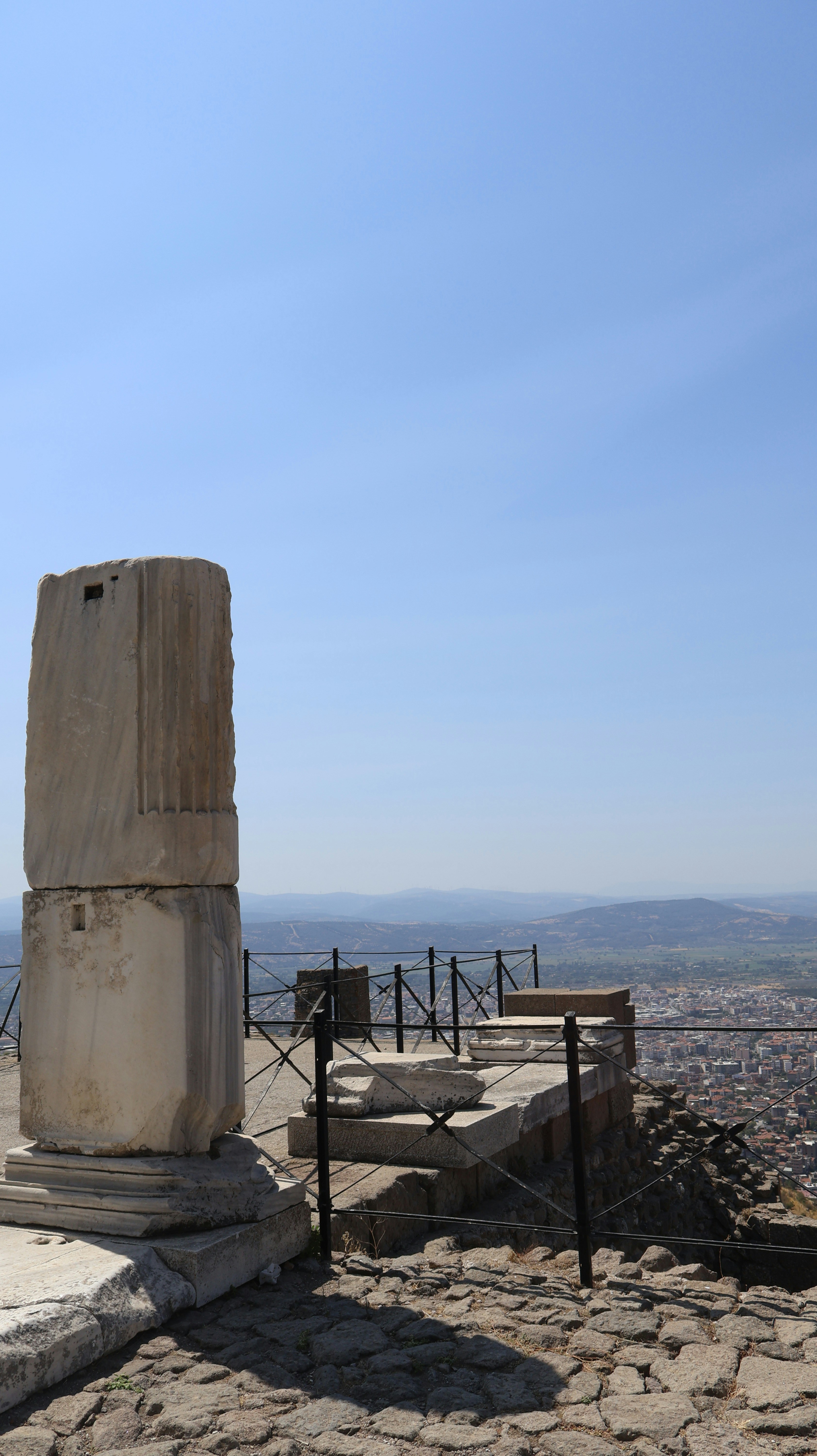 Ancient stone pillar stands against a backdrop of rolling hills and a clear blue sky, overlooking a sprawling city below.