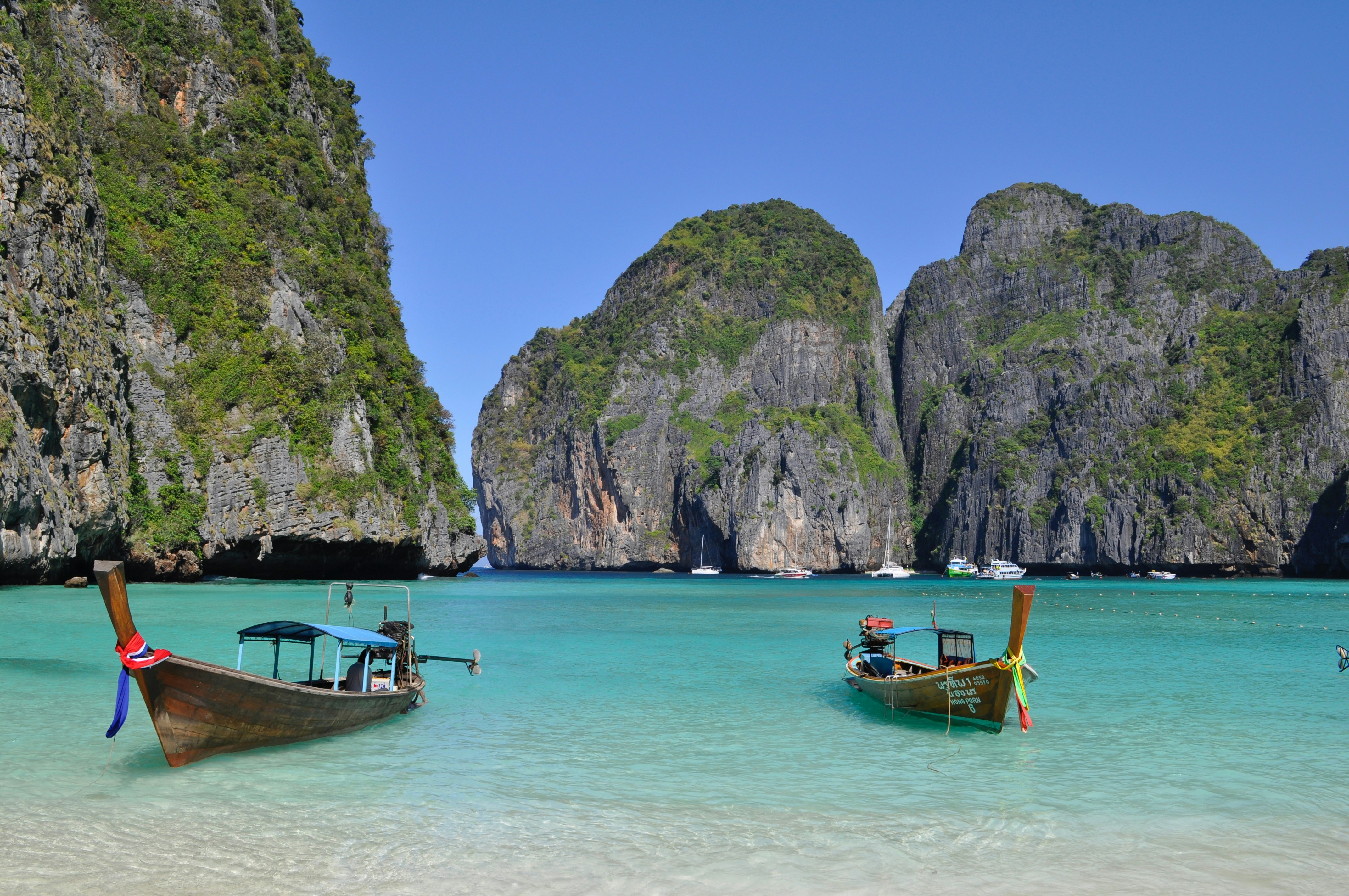 Two longtail boats float in clear turquoise water near cliffs.