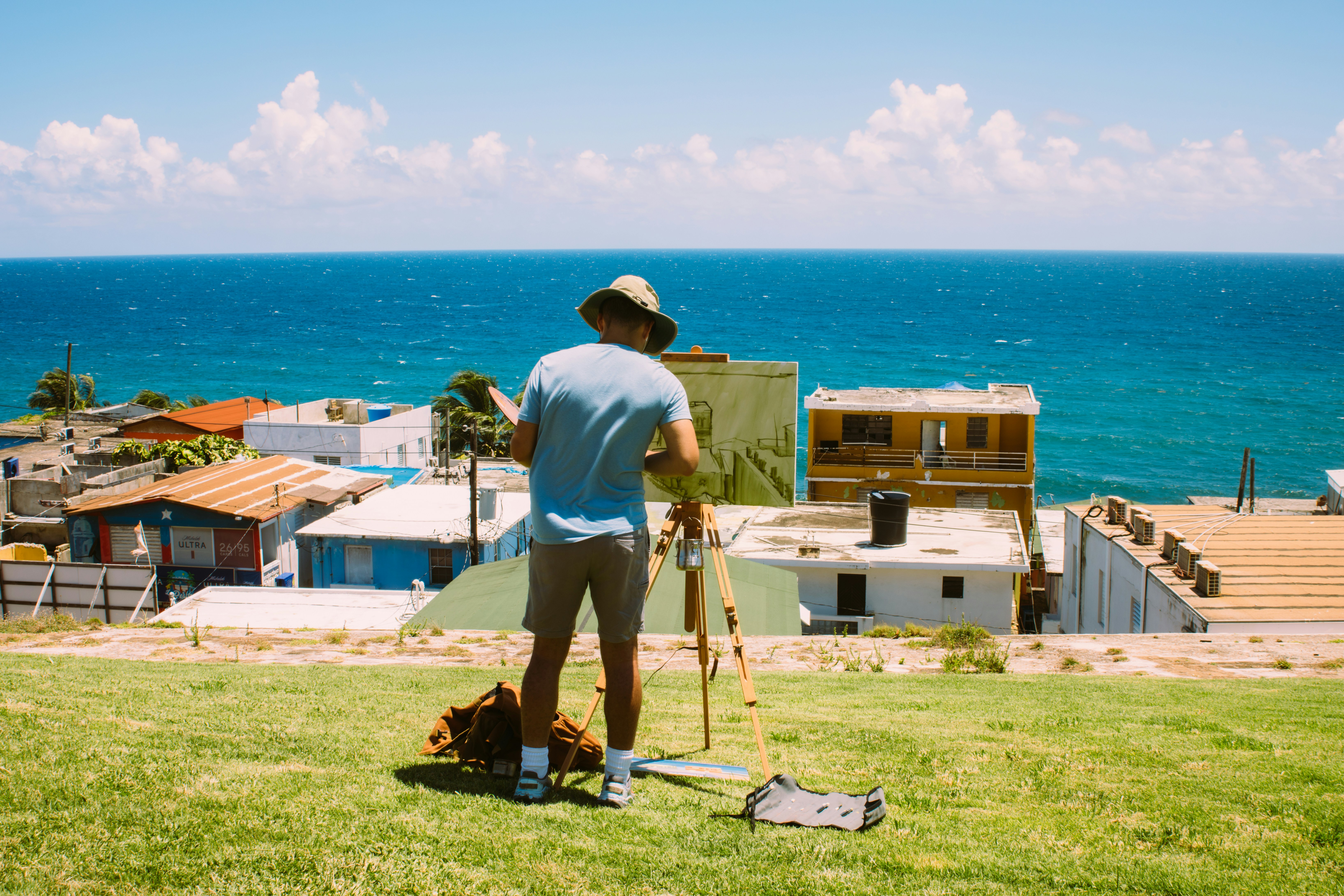 An artist in Old San Juan, Puerto Rico. He is facing the ocean, painting in their outlined canvas. | Artist painting by the sea with colorful houses