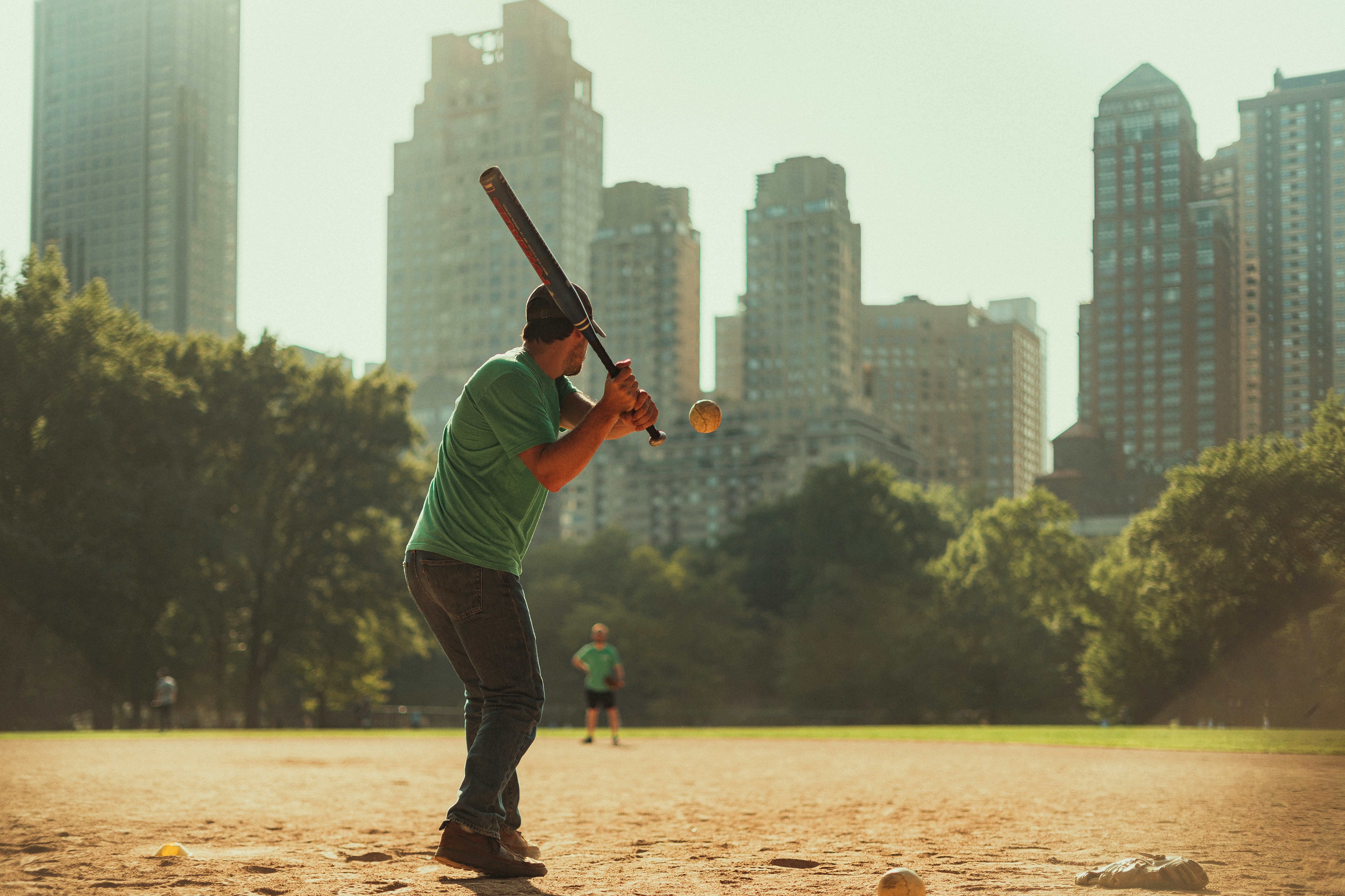 Softball in Central Park | Man swings baseball bat in park with city skyline.