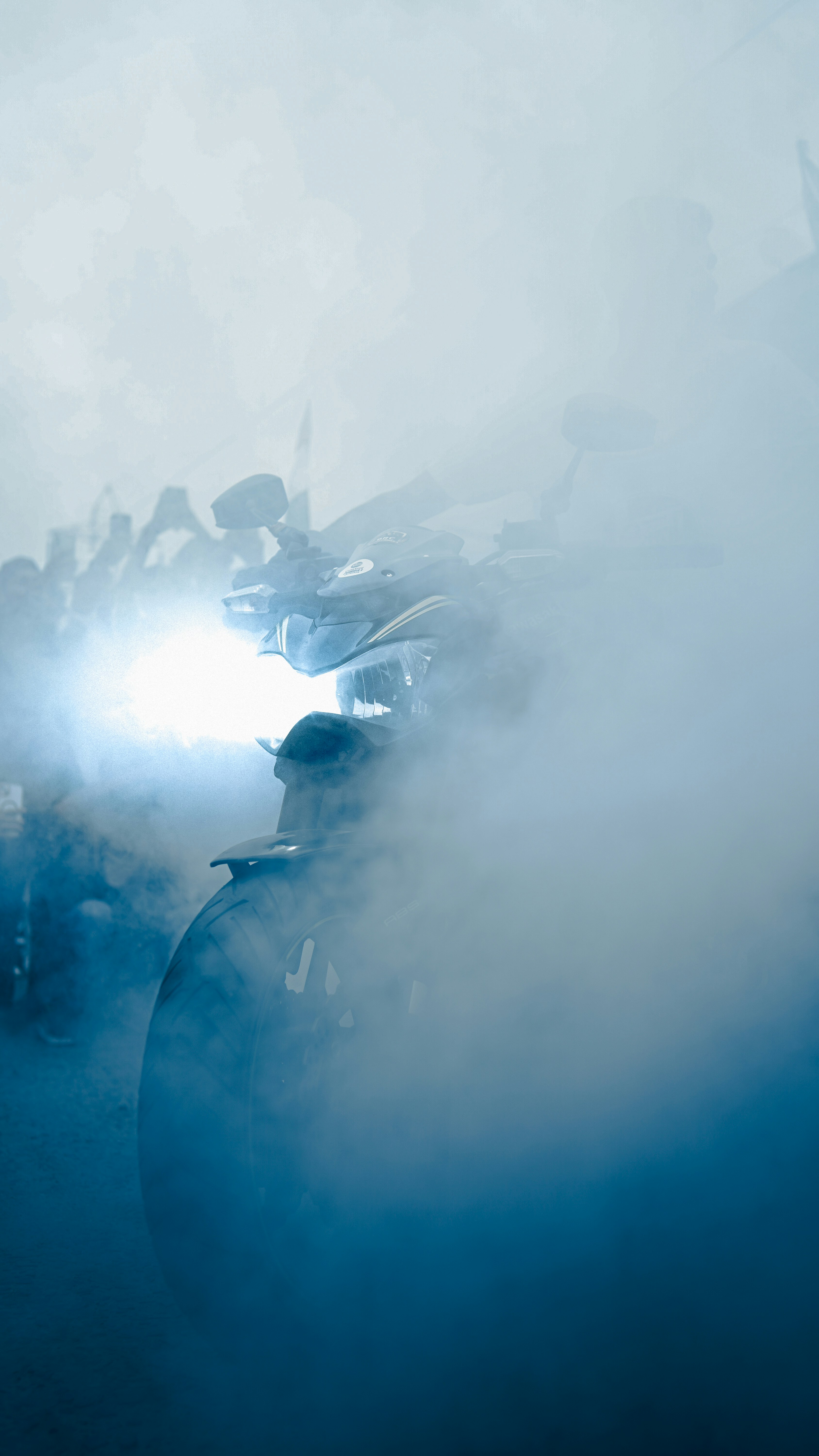 Motorcycle emerging from a dense cloud of smoke, illuminated by bright headlights. The scene captures the essence of speed and excitement.