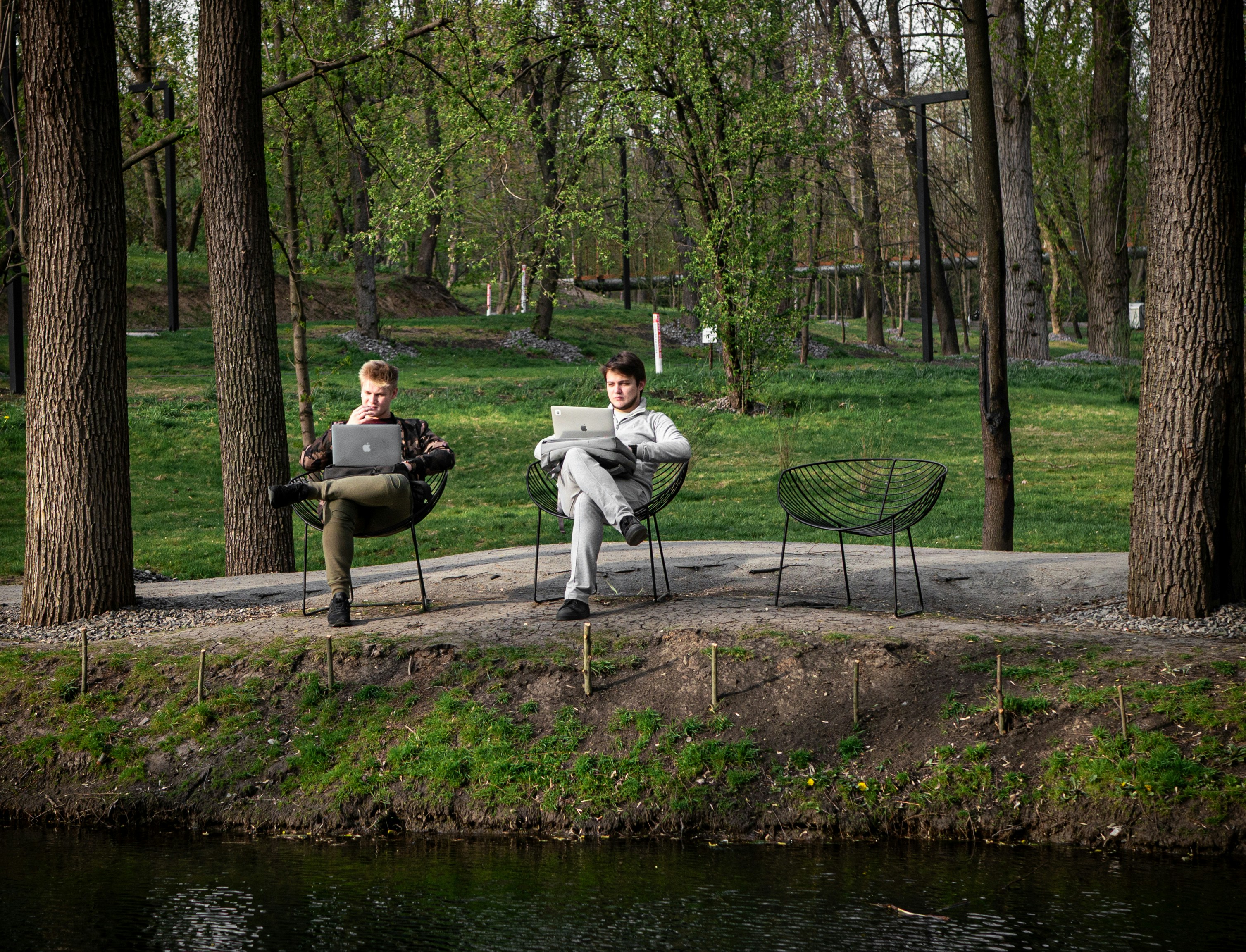 Two people working remotely on laptops while sitting near a quiet park creek. Spring light, trees, and modern chairs frame a peaceful yet paradoxical moment of digital life in a natural setting. | Two men working on laptops in a park