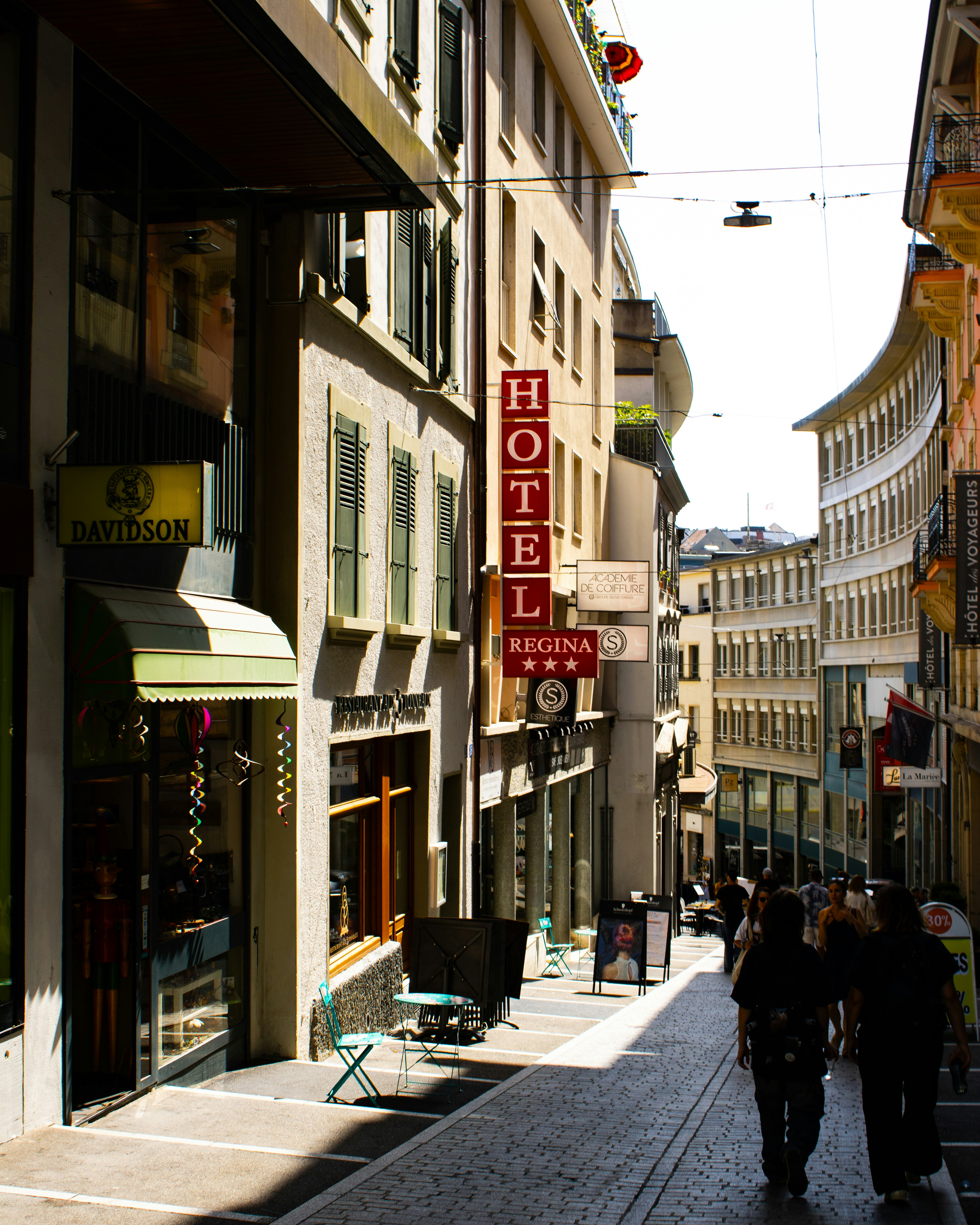 People walk down a narrow street with shops and a hotel.