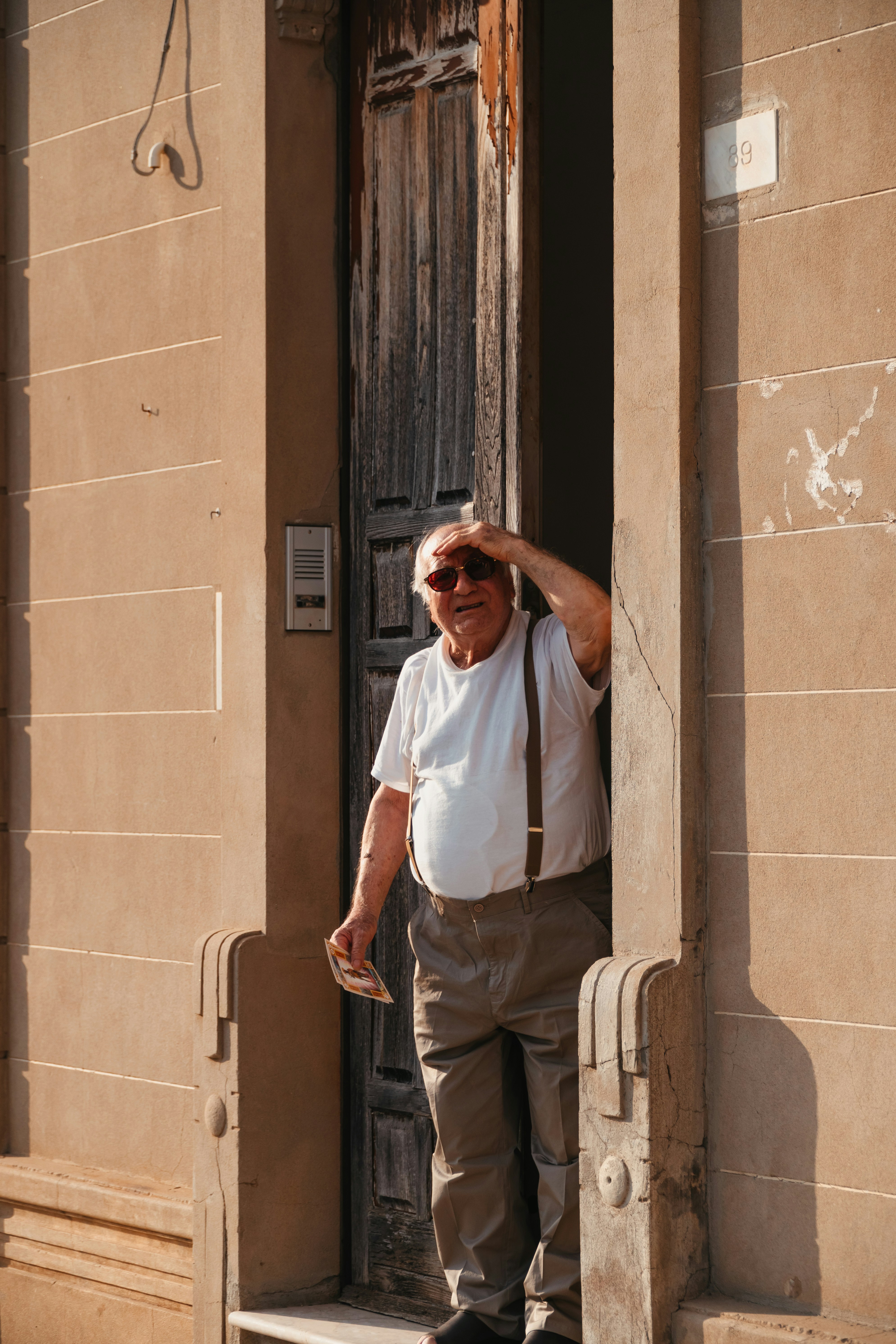 Elderly man in suspenders standing in doorway, shielding his eyes from the sun while holding a brochure.