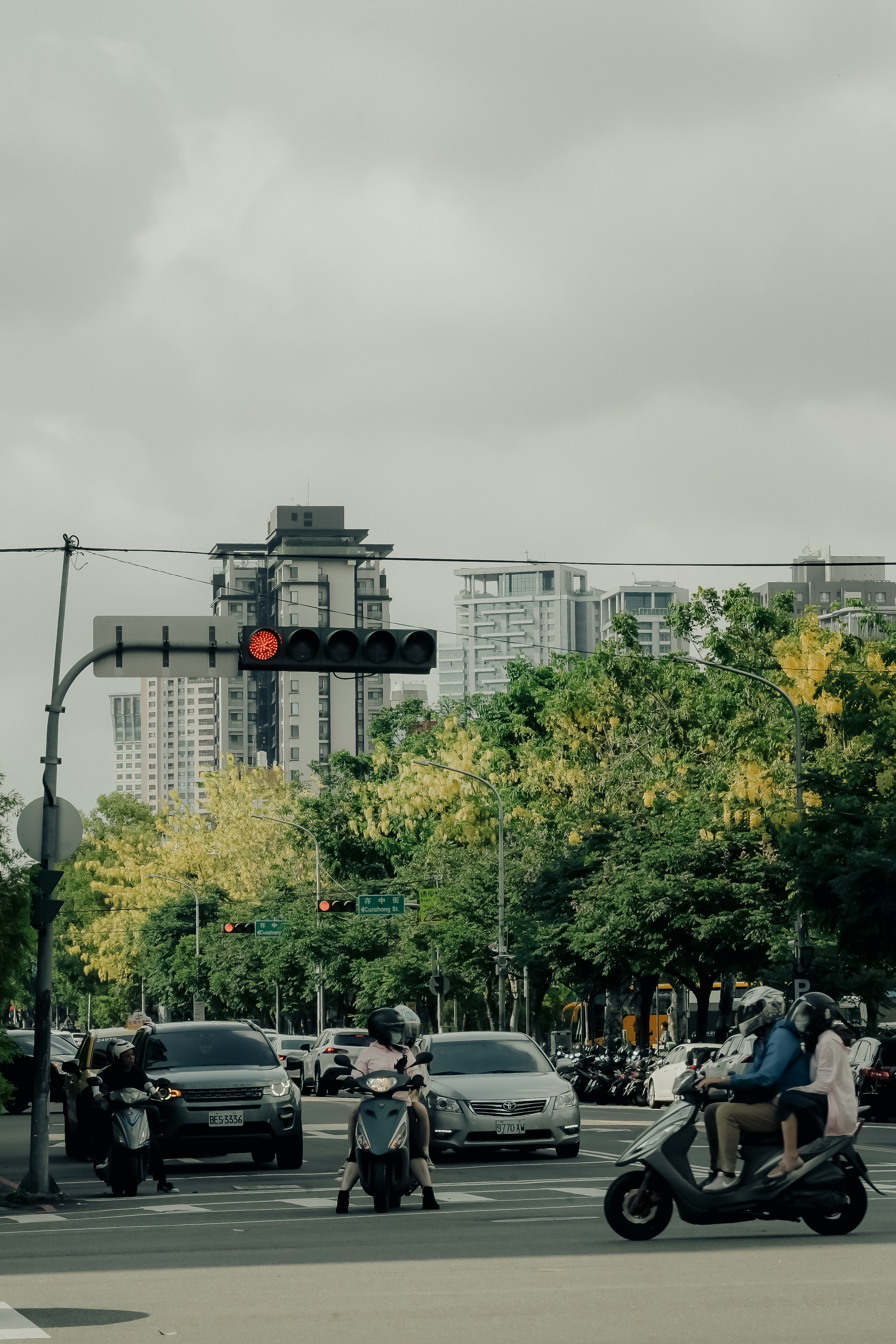 Traffic scene at an intersection featuring vehicles and motorbikes waiting at a red light, framed by city buildings and trees.