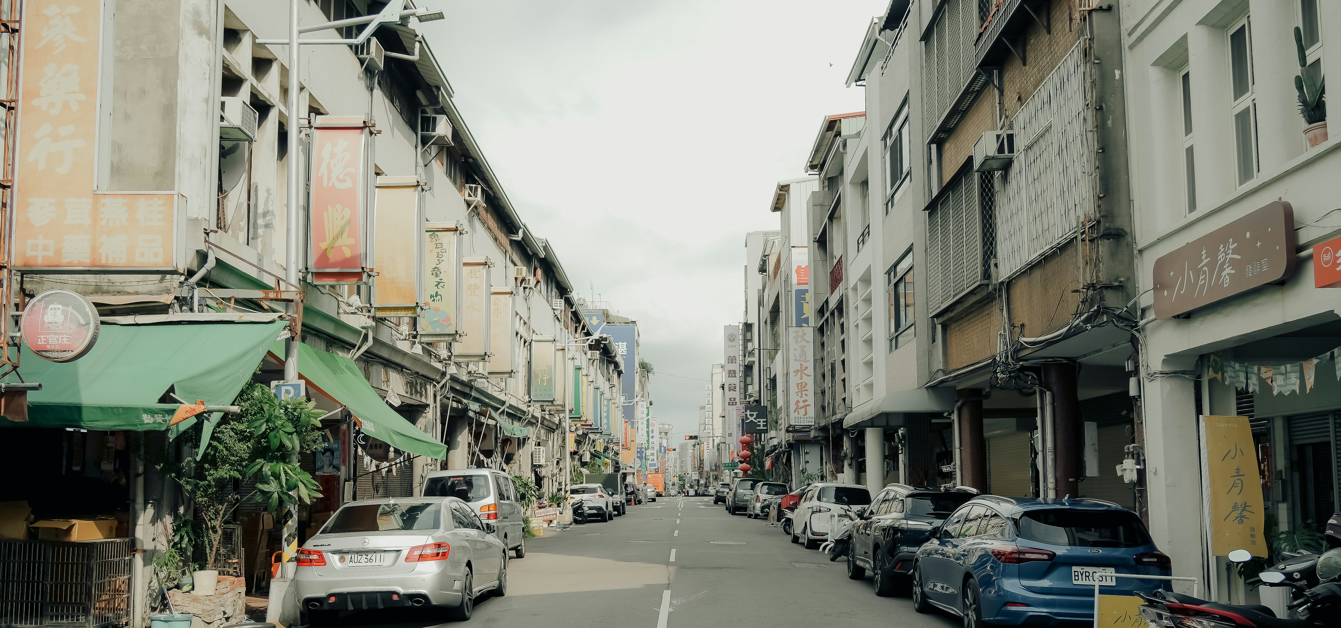 A quiet street lined with vintage buildings and parked cars, showcasing a blend of modern and traditional architecture. The overcast sky adds a moody ambiance.