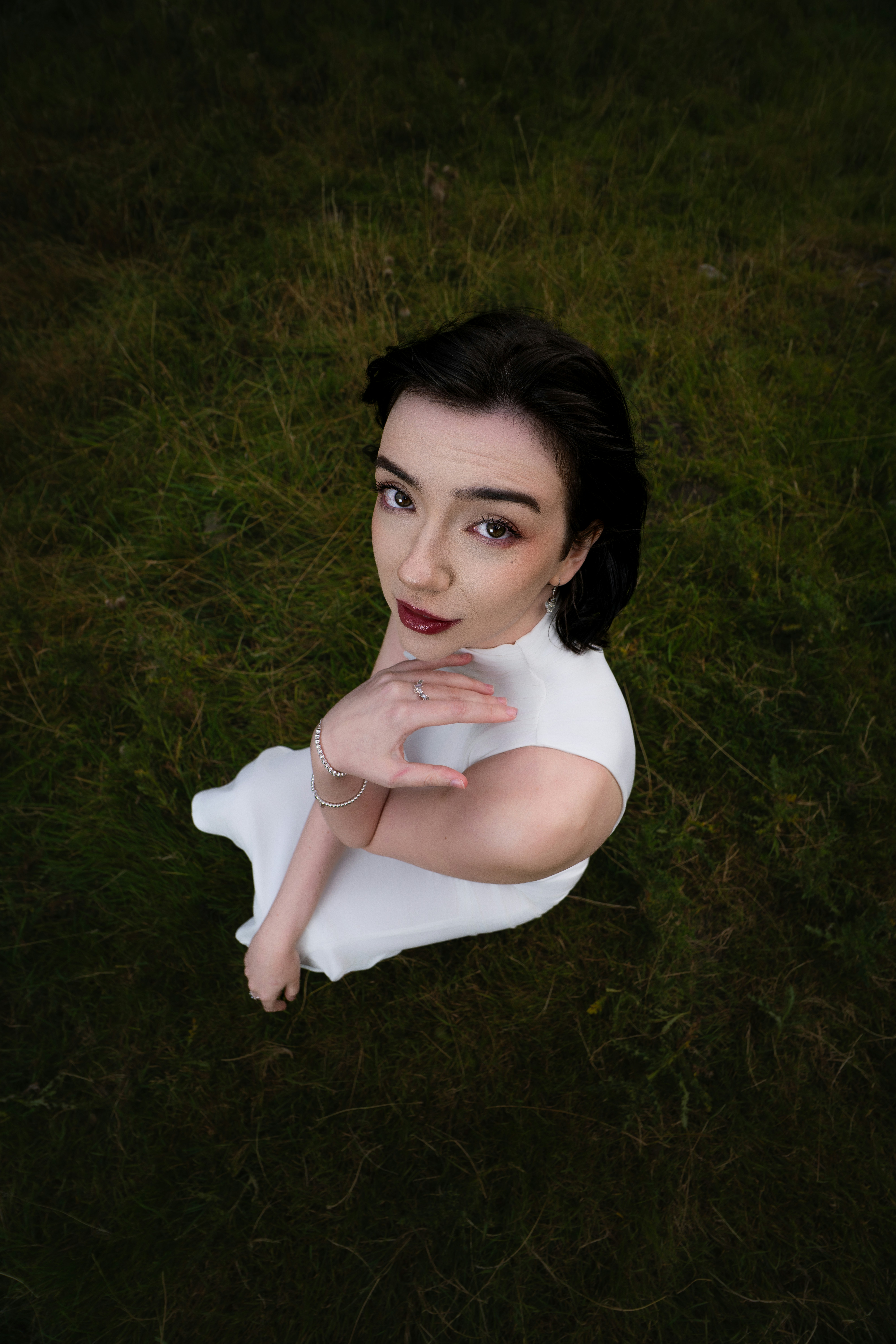 A woman in a white dress gazes upward, seated on lush green grass, exuding a serene and contemplative aura.