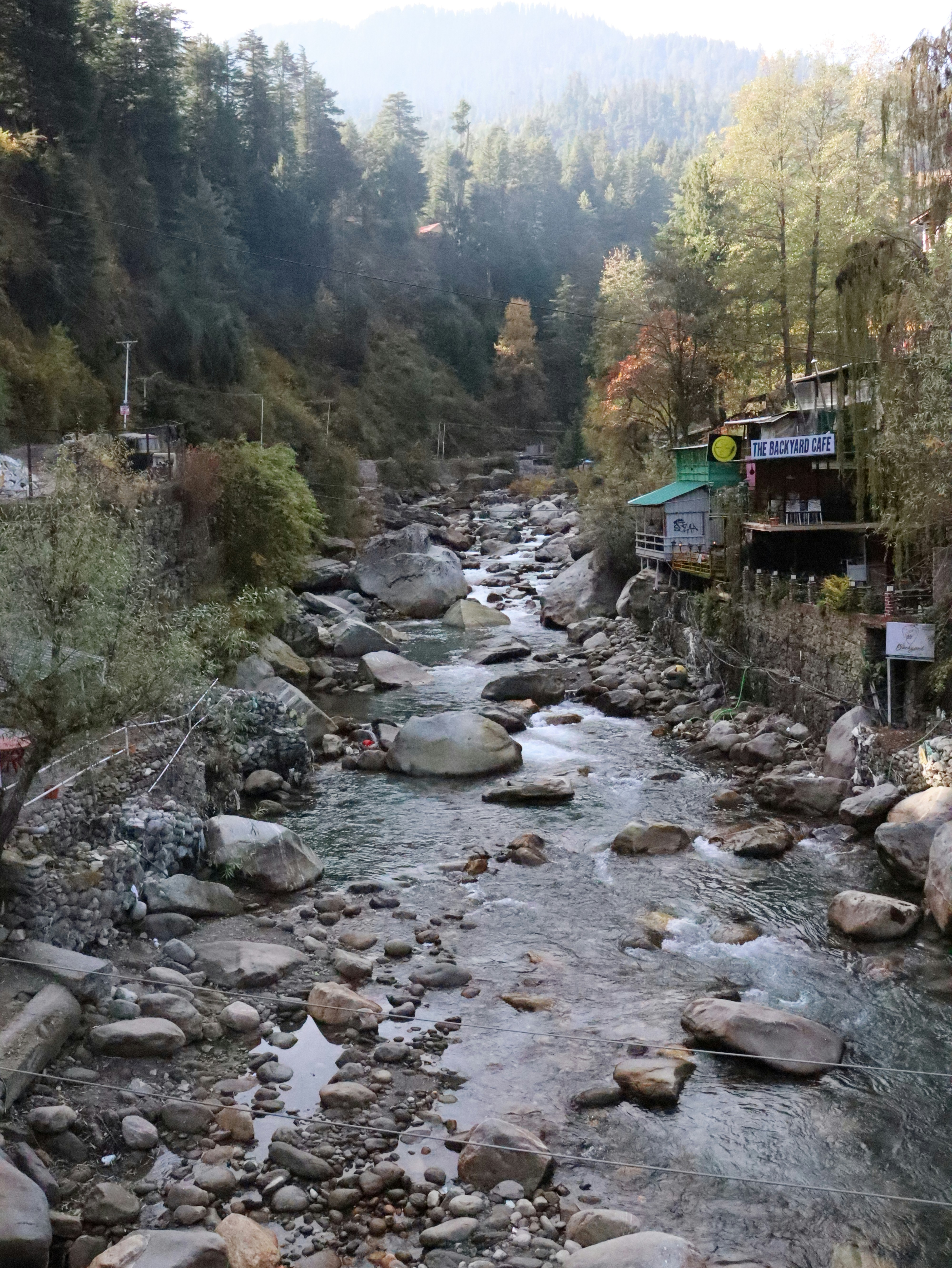 A clear mountain stream winds through a pine-clad valley in Himachal Pradesh, slipping over smooth boulders past rustic riverside cafés. Soft hillside light and forest hues capture the calm energy of the Himalayas. | Rocky river flowing through a forested mountain valley.