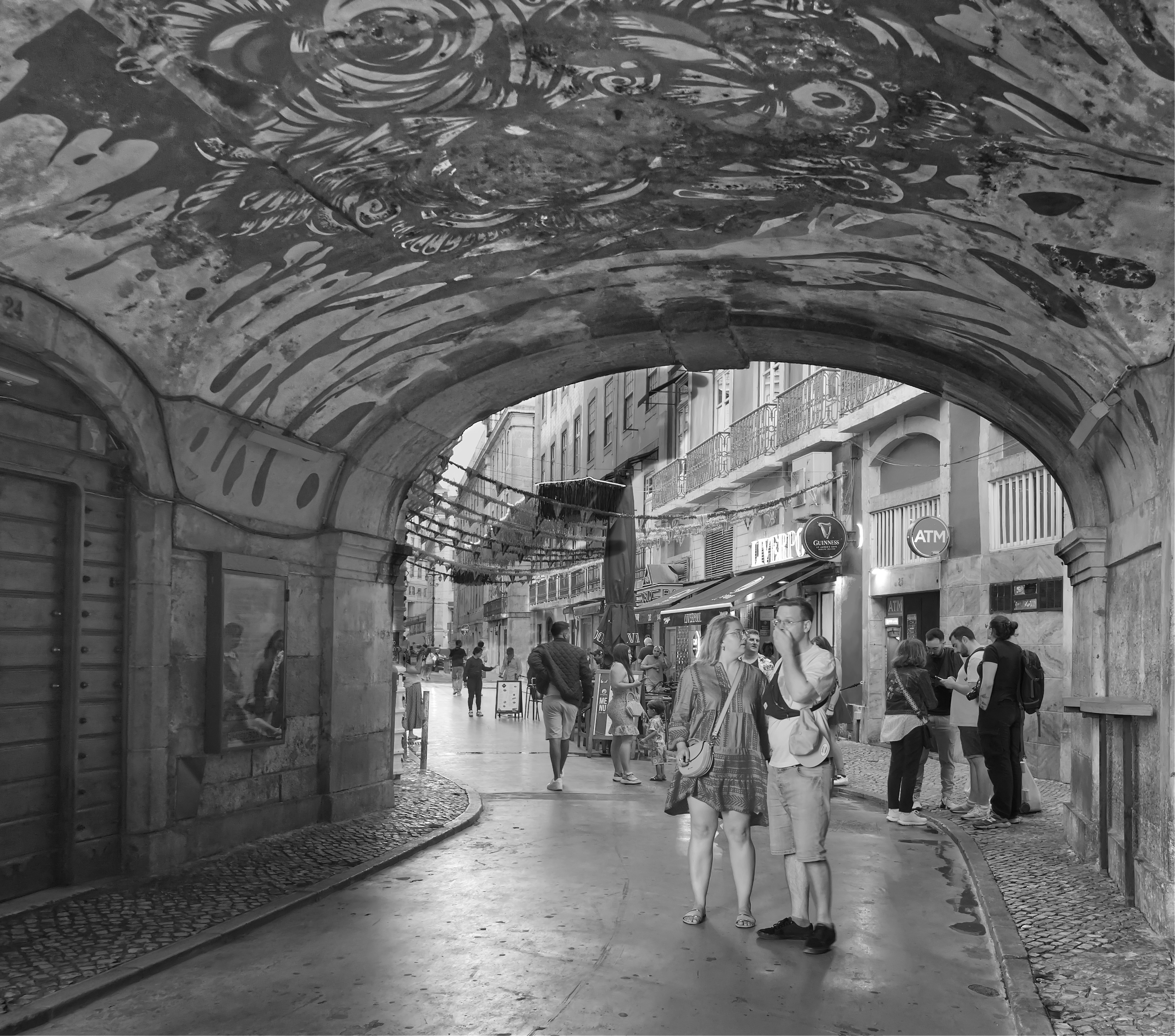 Two tourists pause under an ornate archway adorned with vibrant street art, while locals navigate the bustling street beyond. 