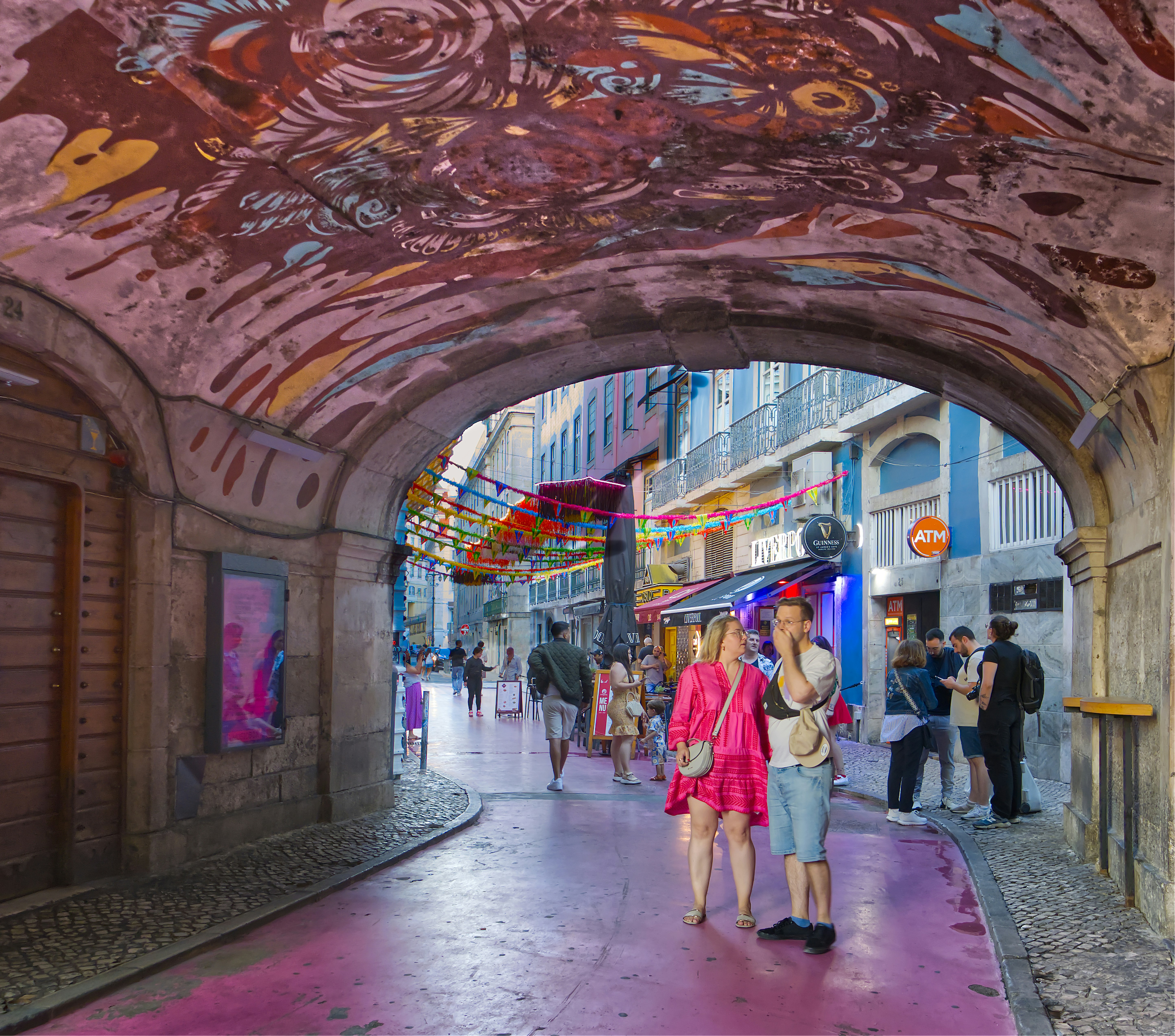 Imaginez une rue étroite, pavée de souvenirs et de secrets, nichée au cœur de Lisbonne. Au début du XXe siècle, la Rua Nova do Carvalho n’était pas le lieu coloré et animé que l’on connaît aujourd’hui. Elle était le repaire des marins en escale, des ombres qui se glissaient entre les maisons closes et les bars enfumés, des tripots où l’on jouait bien plus que de l’argent. C’était un quartier chaud, un peu sulfureux, où la nuit ne finissait jamais vraiment, où les rires se mêlaient aux chuchotements et où l’on préférait ne pas poser trop de questions. En 2011, les pavés de la rue se sont parés d’une teinte rose vif, presque provocante, comme un clin d’œil à son histoire tumultueuse. Aujourd’hui, la rue Rose est un lieu où l’on vient pour faire la fête. Dès que le soleil commence à descendre, les terrasses s’animent, les verres s’entrechoquent, et les rires résonnent entre les façades colorées. Les bars branchés, les restaurants conviviaux et les clubs musicaux ont remplacé les anciennes enseignes douteuses. | Colorful street art adorns arched walkway with people