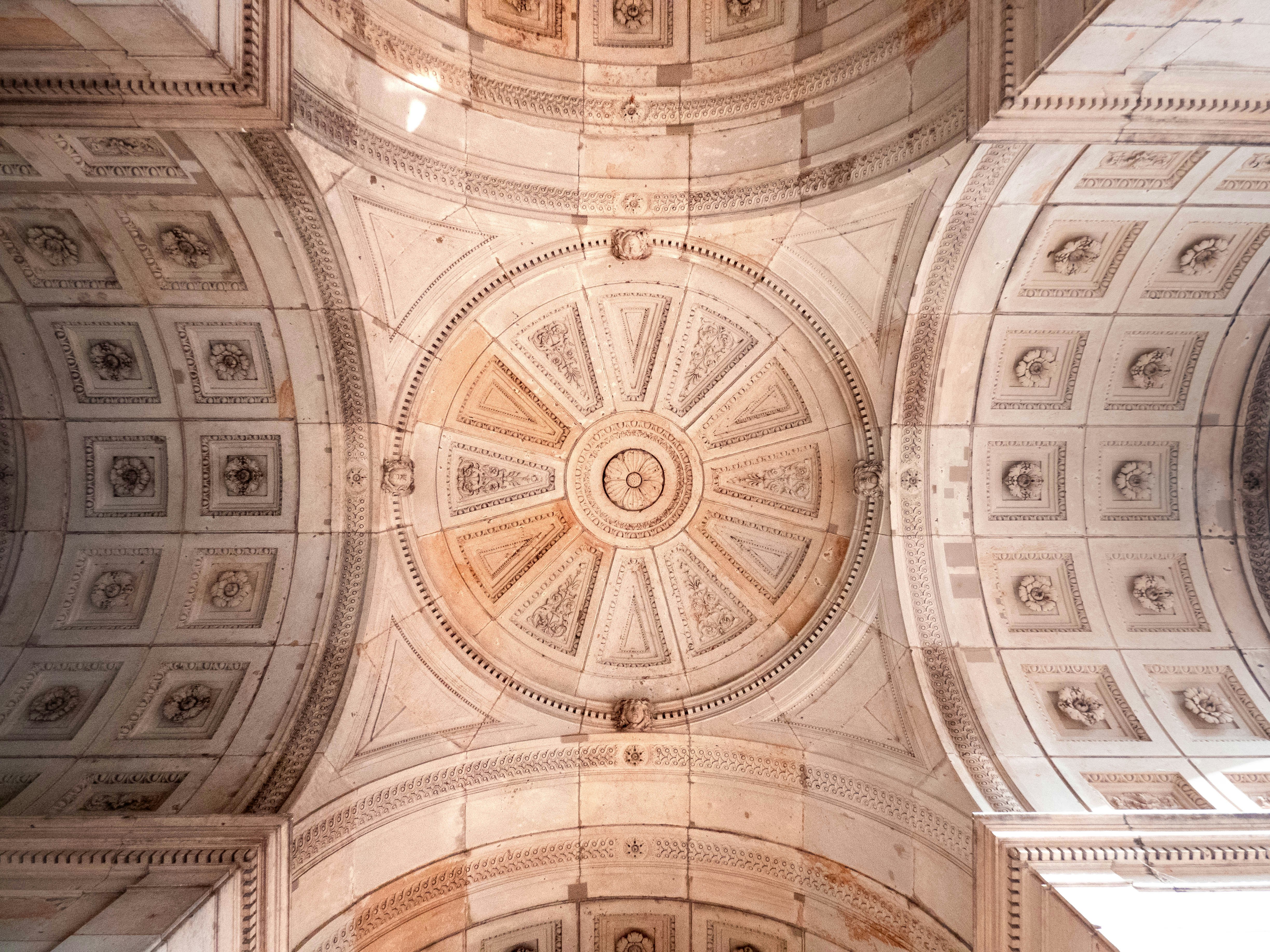 Ceiling view of a stone dome with decorative geometric patterns and arches, featuring classical architectural elements and symmetrical ornamentation. | Intricate stone ceiling with circular and square patterns