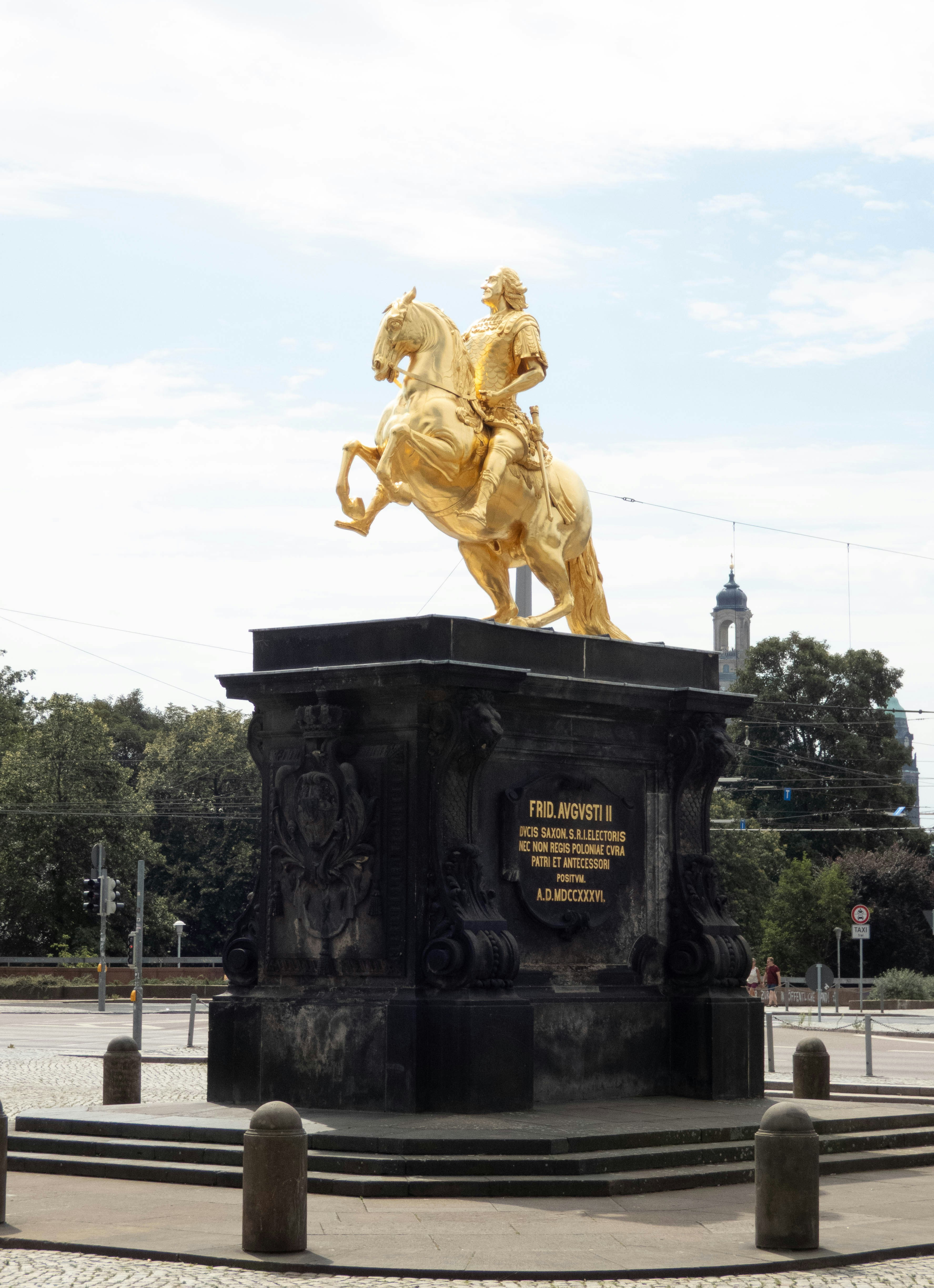 A golden equestrian statue stands prominently on a dark stone pedestal, depicting a rider in armor. The monument is located outdoors in an urban square with surrounding trees and historic buildings visible in the background. The pedestal features an inscription with the name "FRID. AUGUSTII II" and details about the monument. | Golden equestrian statue on a dark pedestal