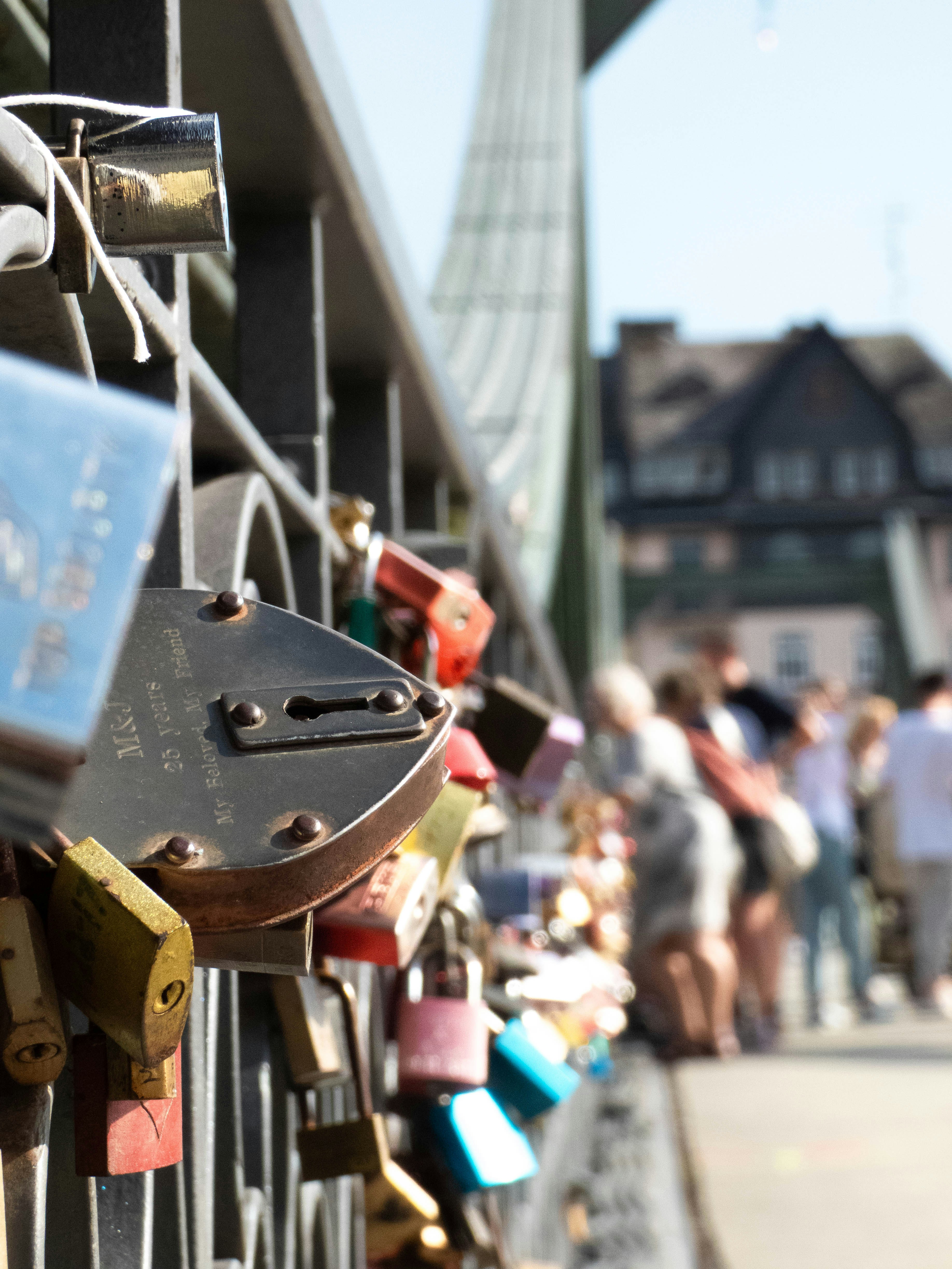 Padlocks attached to the metal railing of a bridge in an urban environment in Frankfurt, Germany. The locks are of different shapes and colors. In the background, there are blurred figures of people walking and buildings with pitched roofs. The photo is taken during daylight. | Love locks attached to a bridge railing with people in background
