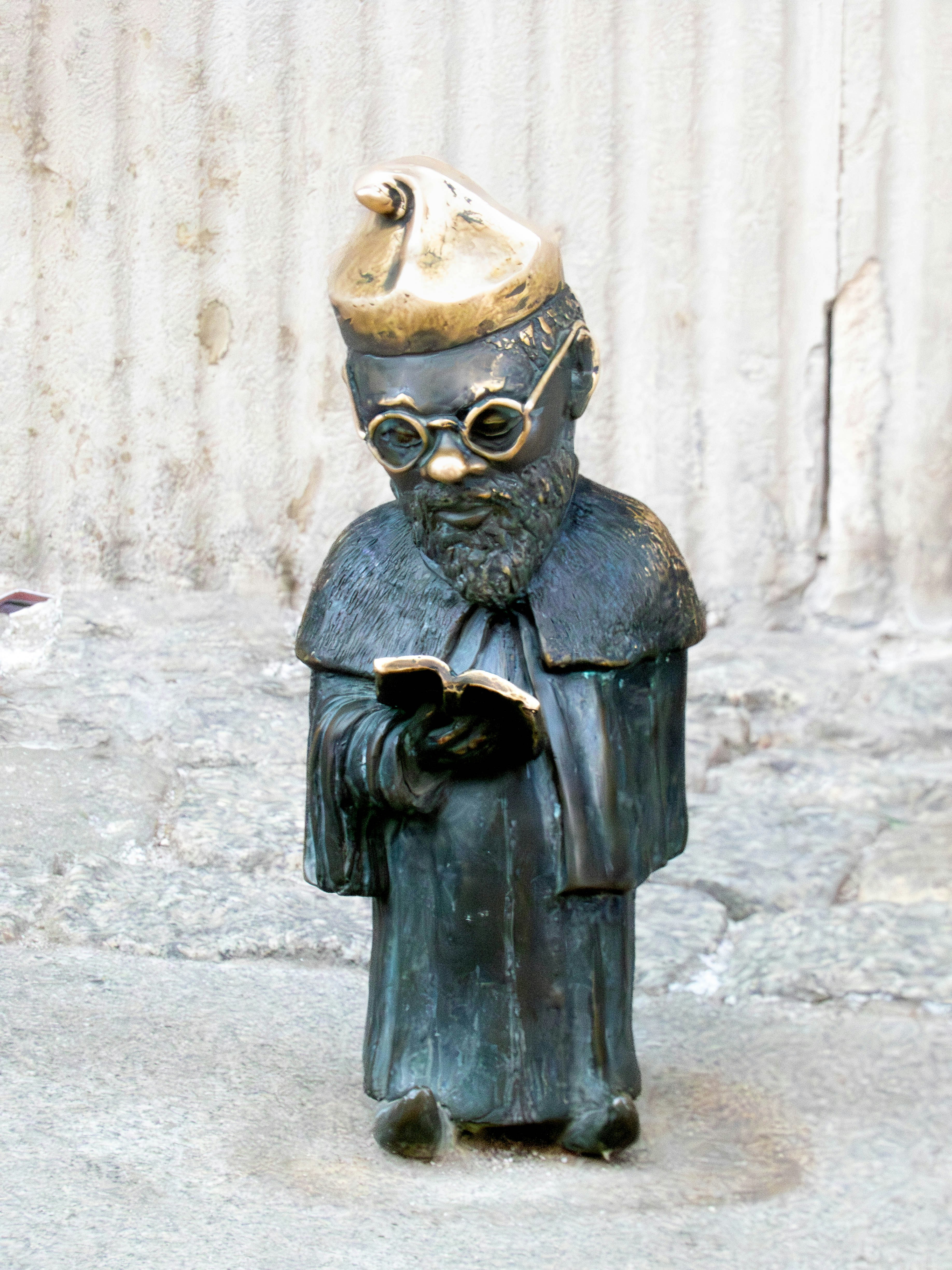 A small bronze statue of a dwarf in a robe holding an open book, standing on a paved outdoor surface. The statue features a pointed hat and is part of a series of similar figures placed throughout Wroclaw, Poland. The background consists of a textured wall and stone pavement. | A bronze statue of a dwarf reading a book.