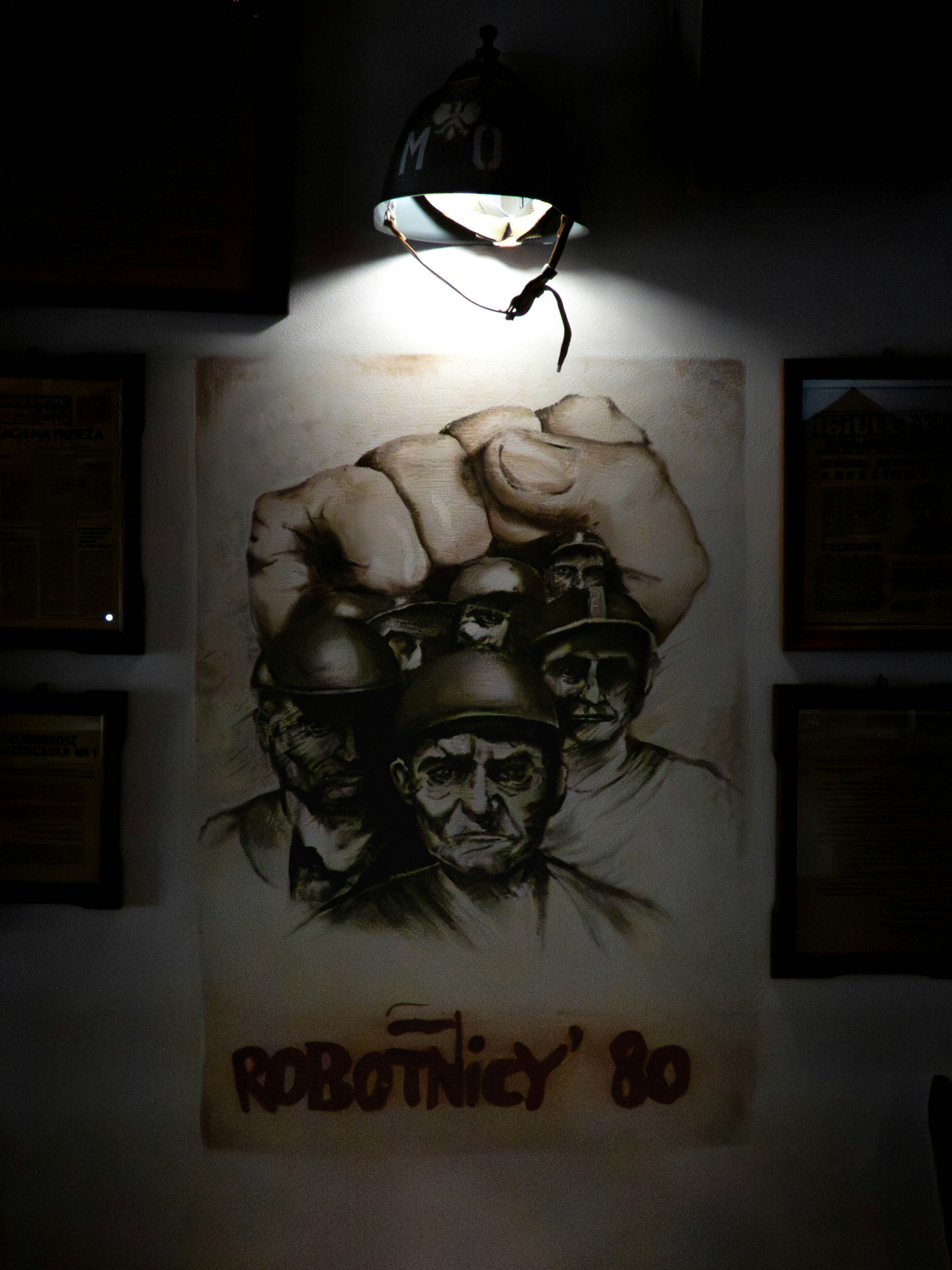 A wall decoration in a restaurant in Wroclaw, Poland, showing a mural of workers wearing helmets with a raised fist in the background. Above the mural, a helmet with the letters "MO" is used as a lamp. The text on the mural reads "Robotnicy '80". | Poster with fist and faces under spotlight