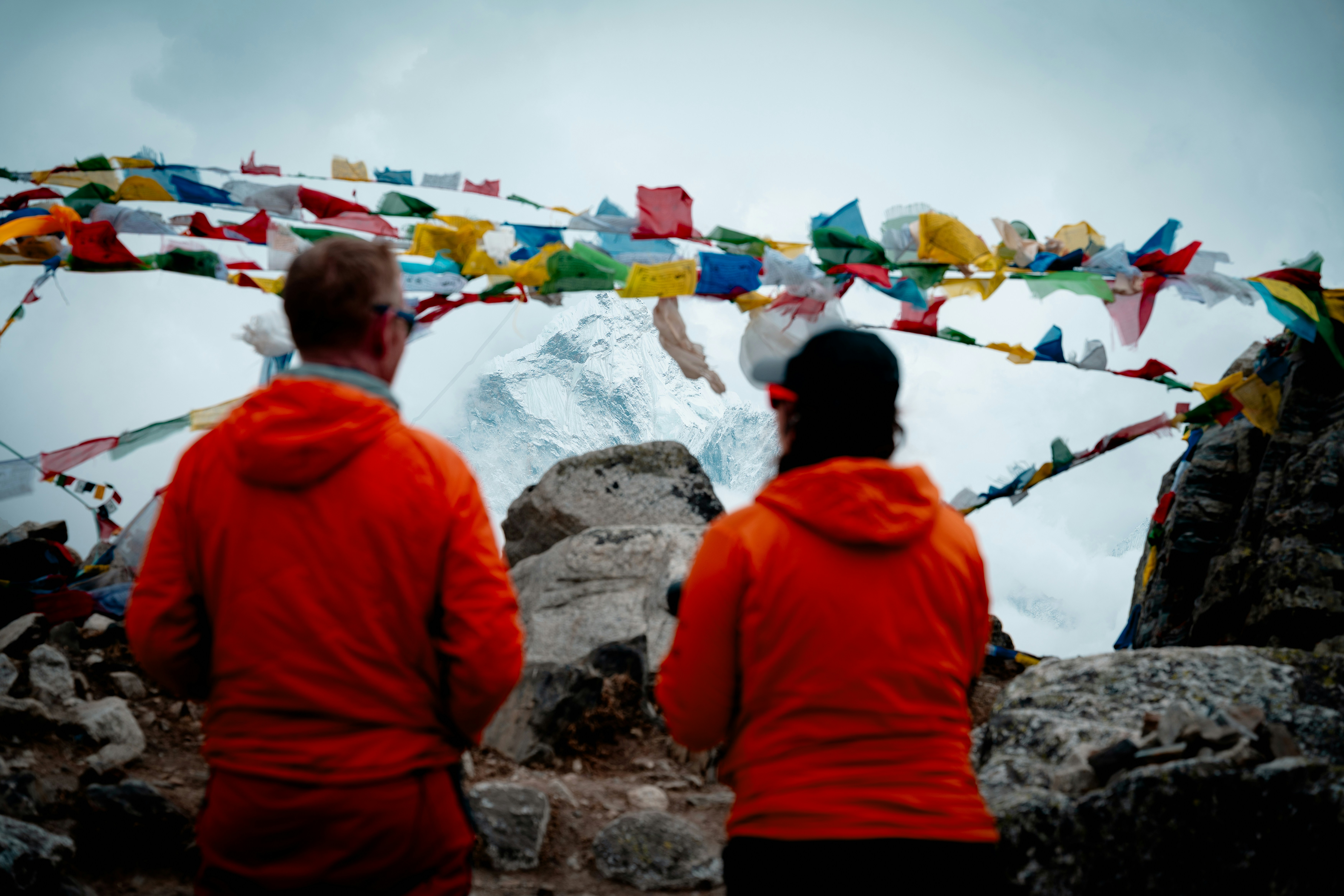 Trekkers enjoying the spectacular sight of Ama Dablam from Thugla Pass, en route to Everest Base Camp Trek. | Two people in orange jackets look at prayer flags