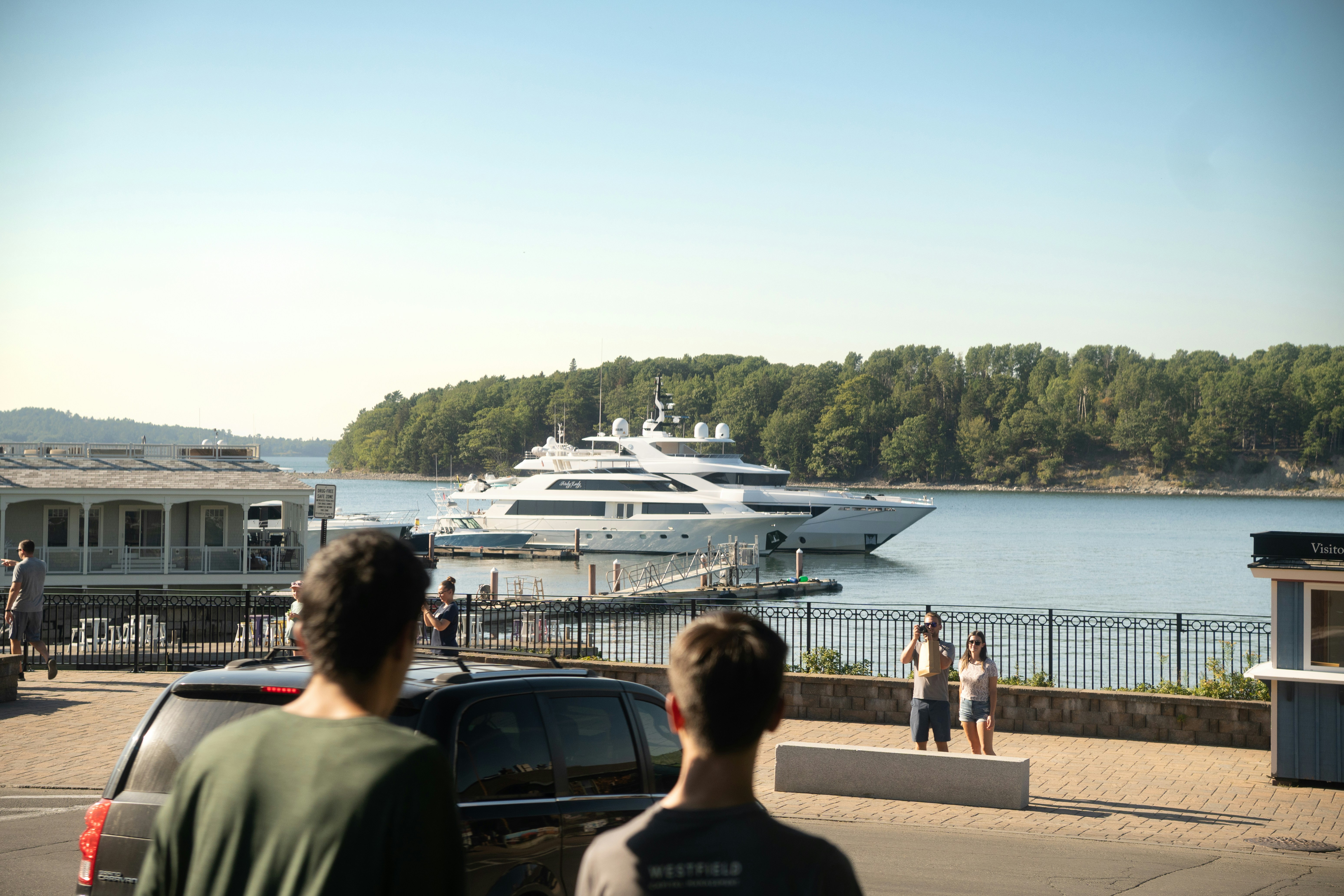 A sleek yacht anchored in a serene bay, with onlookers capturing the moment on their phones. The lush greenery in the background enhances the tranquil atmosphere.