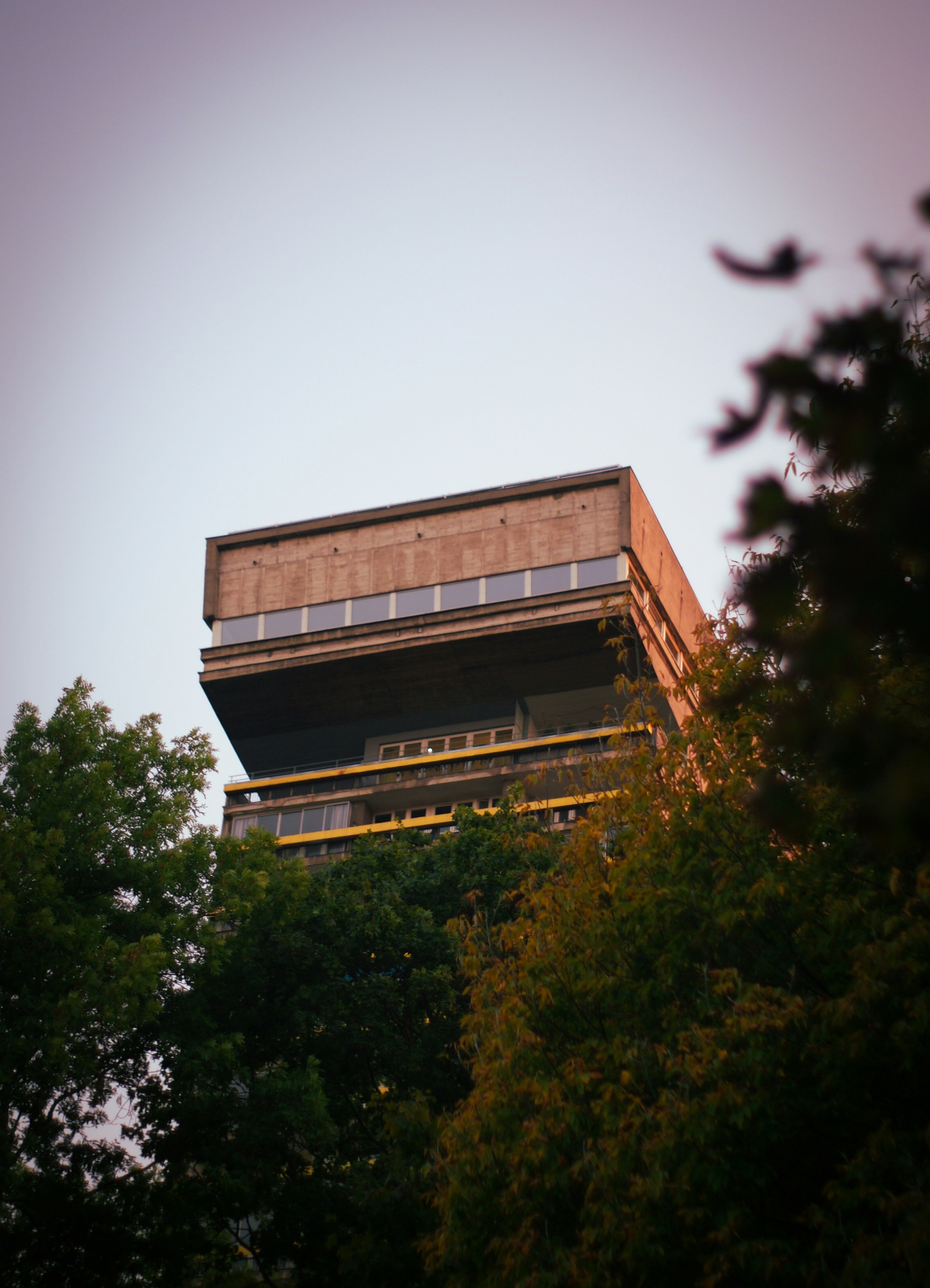 A striking modern building perched above lush greenery, showcasing a unique architectural design against a pastel sky.