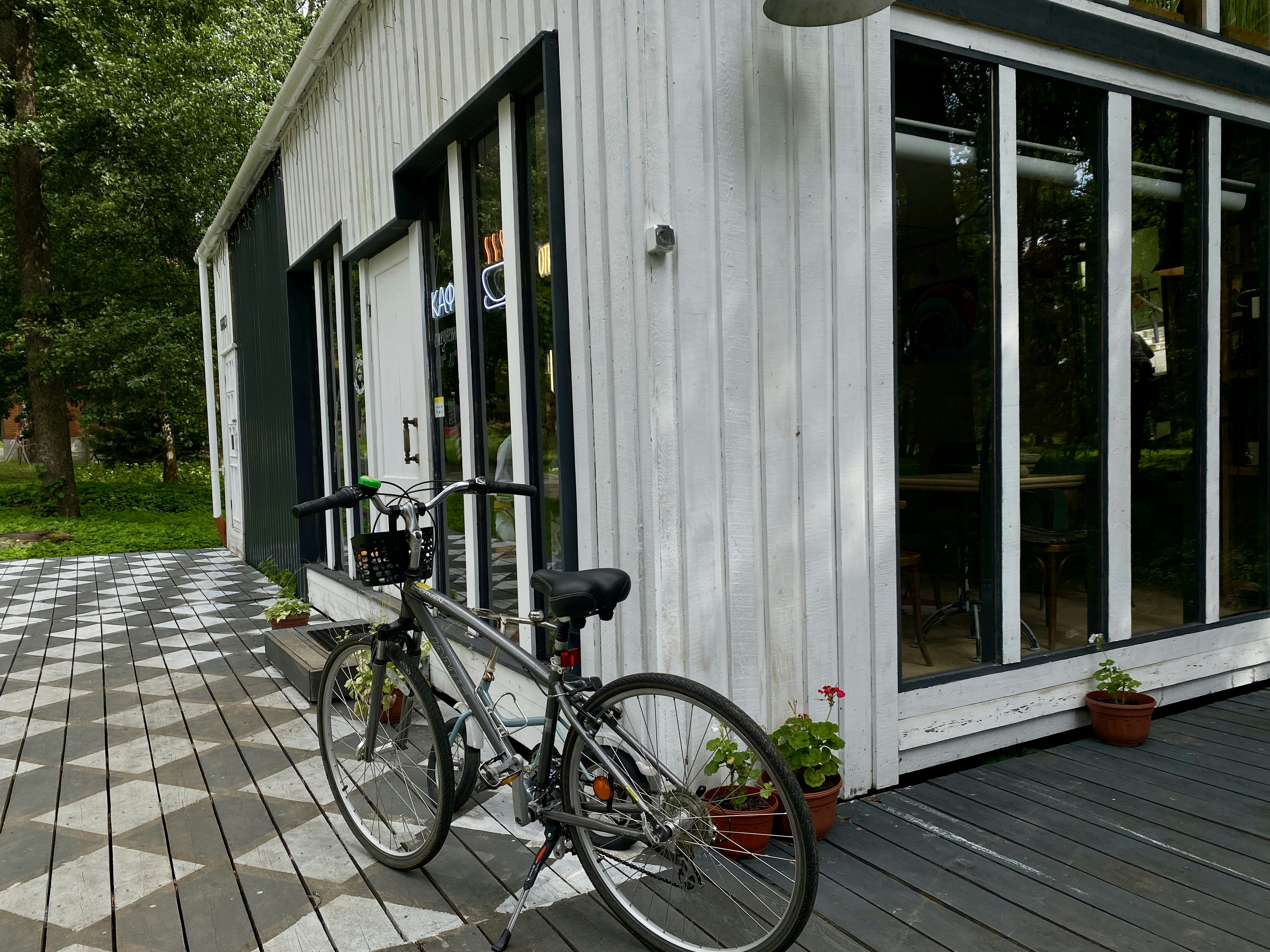 A bicycle parked beside a modern, white wooden structure with large windows, surrounded by greenery and decorative pots. The pattern on the deck adds a playful touch.