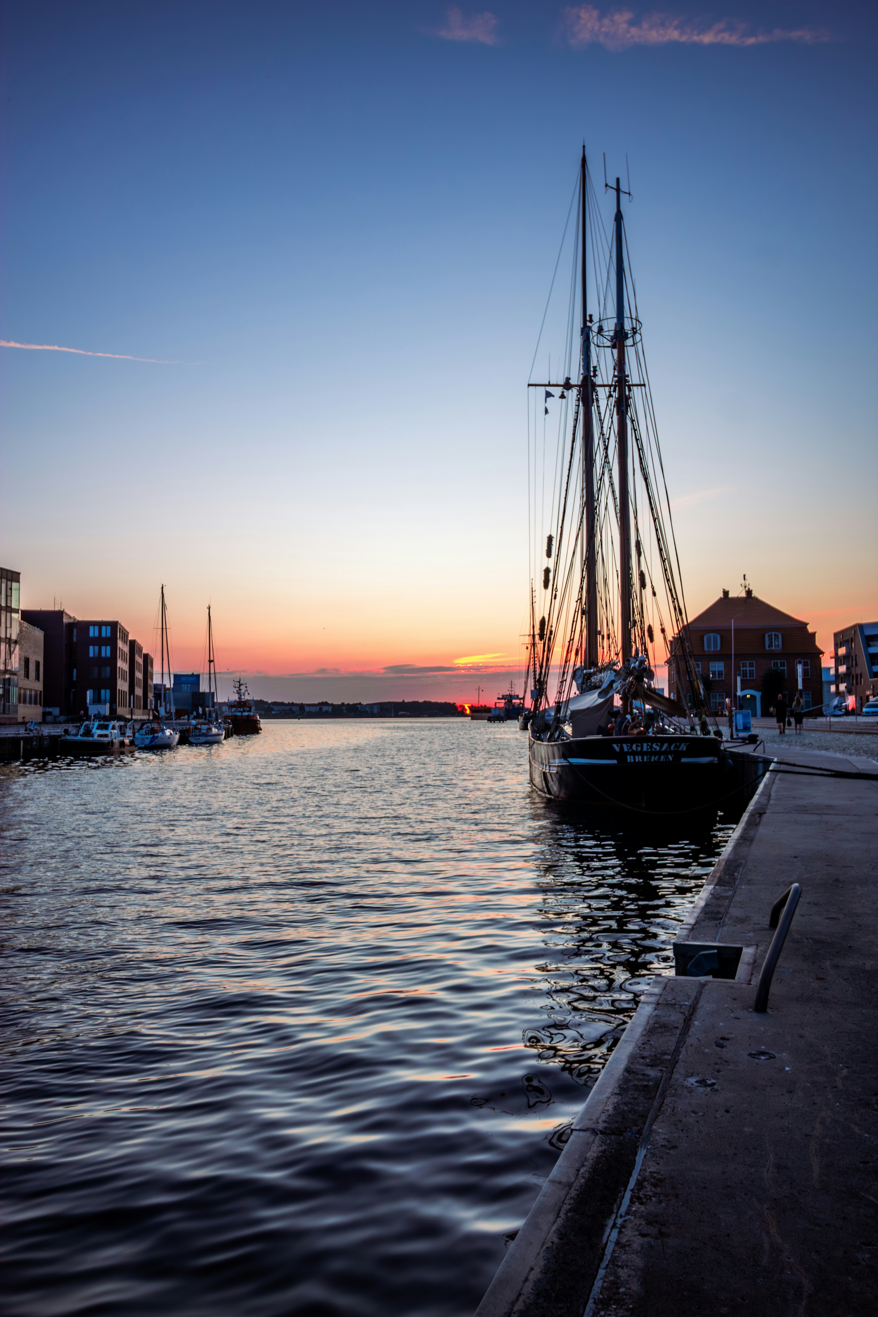 Sailboat docked in harbor at sunset with buildings.