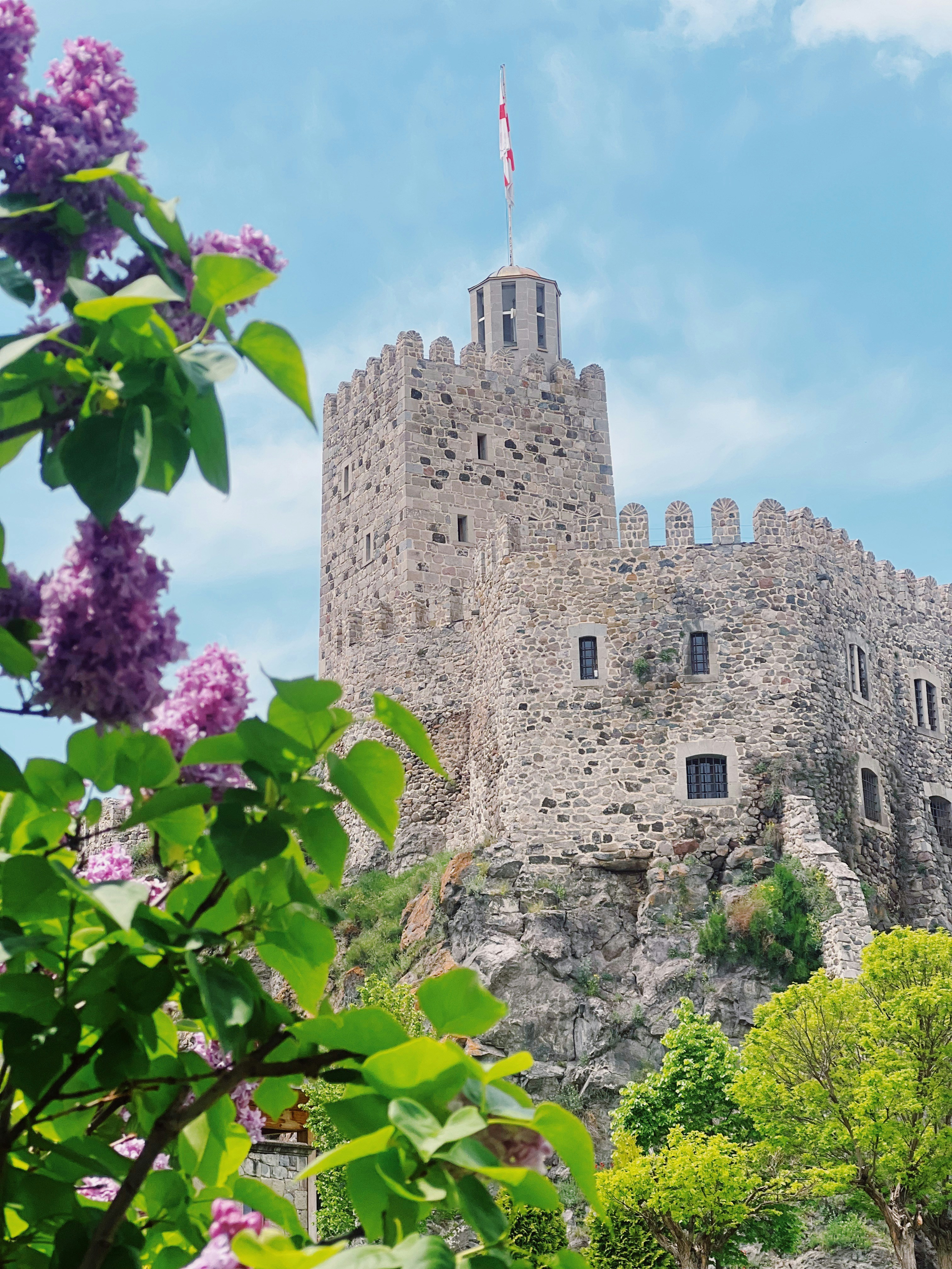 A view of the historic Akhaltsikhe Fortress (Rabati Castle) in Georgia. Travel and architecture photography capturing medieval heritage and Georgian history. | Stone castle with flag on a hill, purple flowers foreground