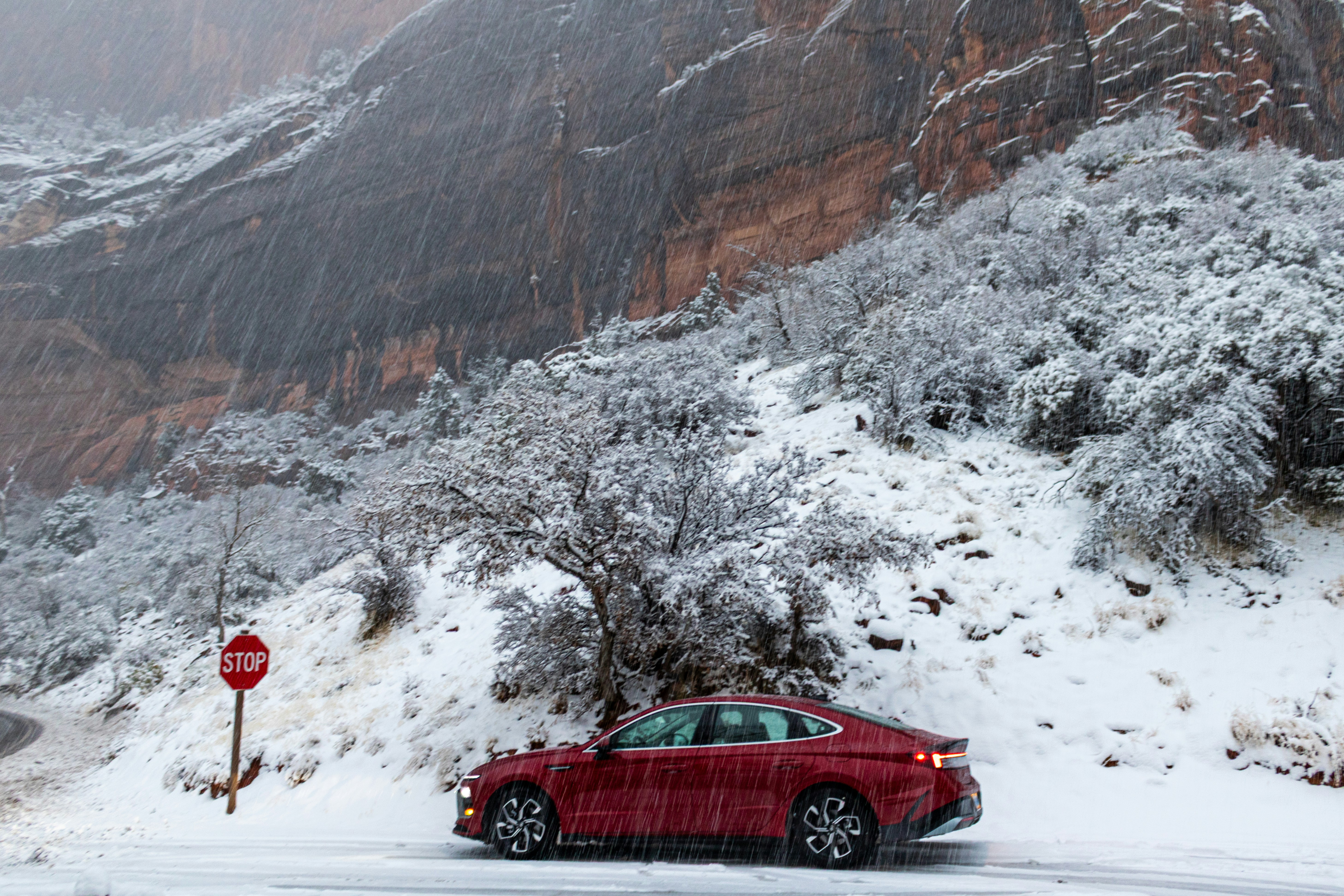 A red Hyundai Sonata parked in the midst of a snowstorm at Zion National Park, Utah. The car’s bold color stands out against the muted whites and grays of a cold winter day, creating a striking visual contrast. | Red car parked on snowy road near red rock cliff.