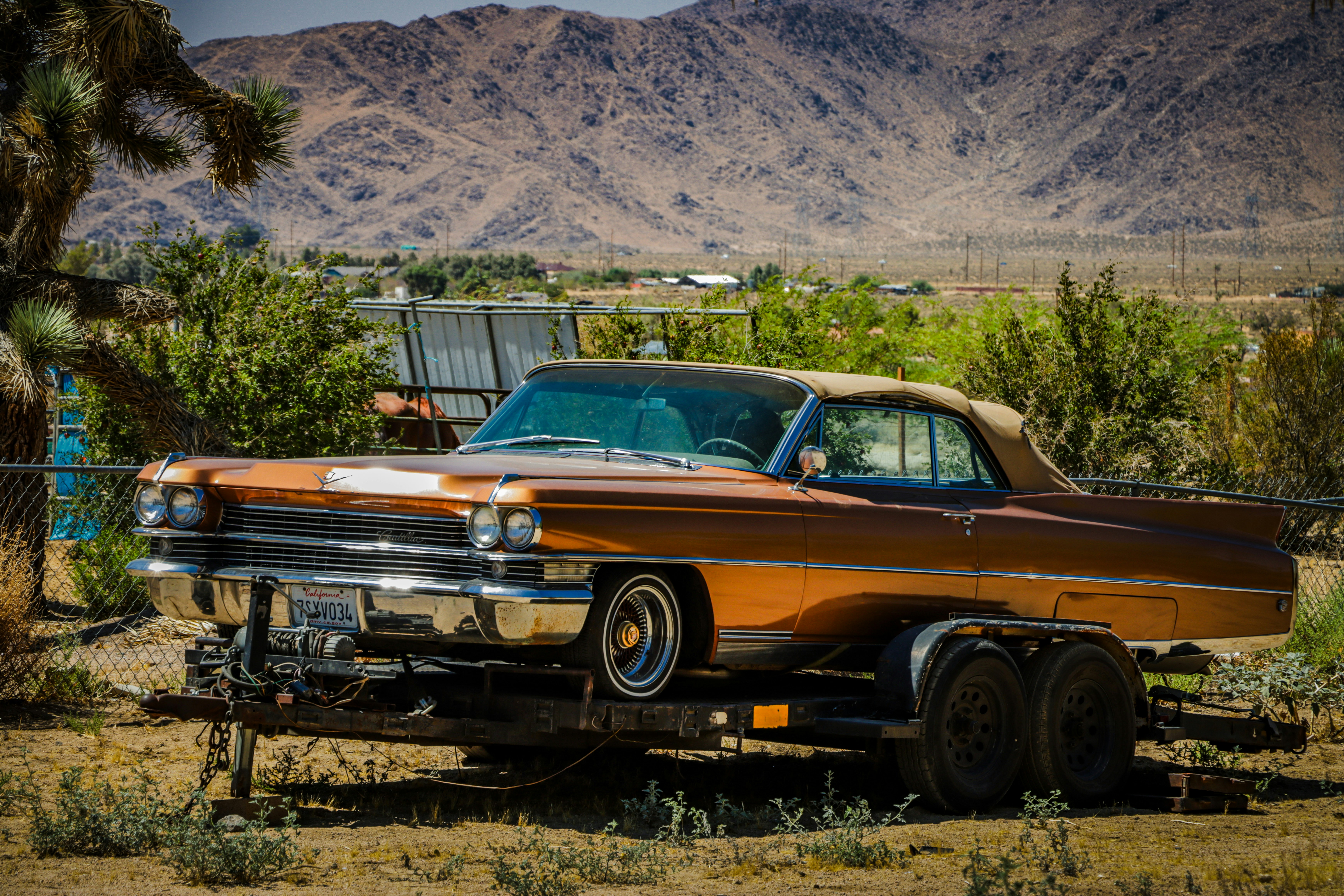 A vintage convertible car rests on a trailer amidst a desert landscape, framed by distant mountains and sparse vegetation.