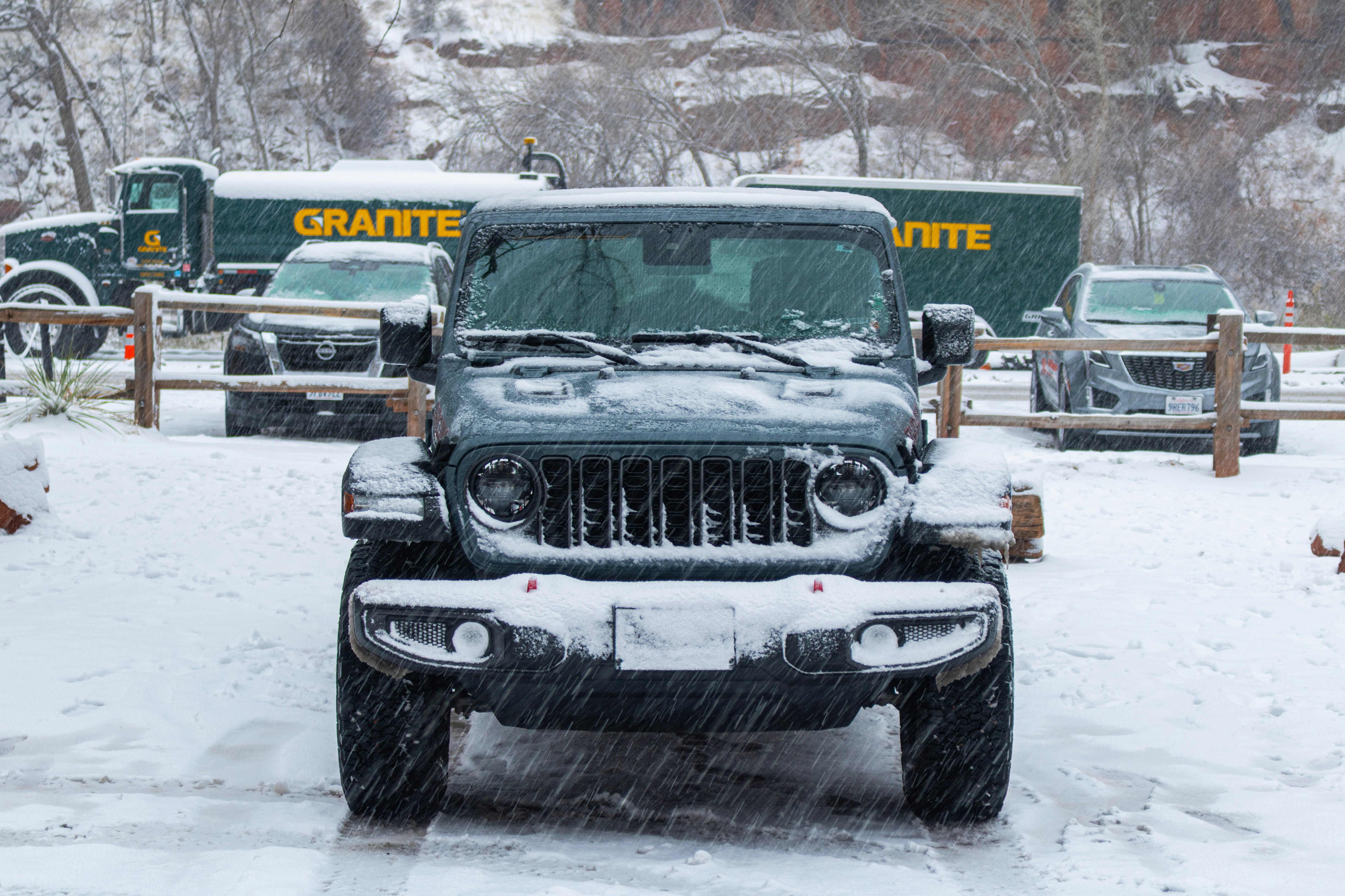 A Jeep Wrangler is parked in the snow filled parking lot of Zion National Park. It is covered in snow dust and stands majestically in the snow storm on cold February winter day. | A dark jeep wrangler parked in the snow