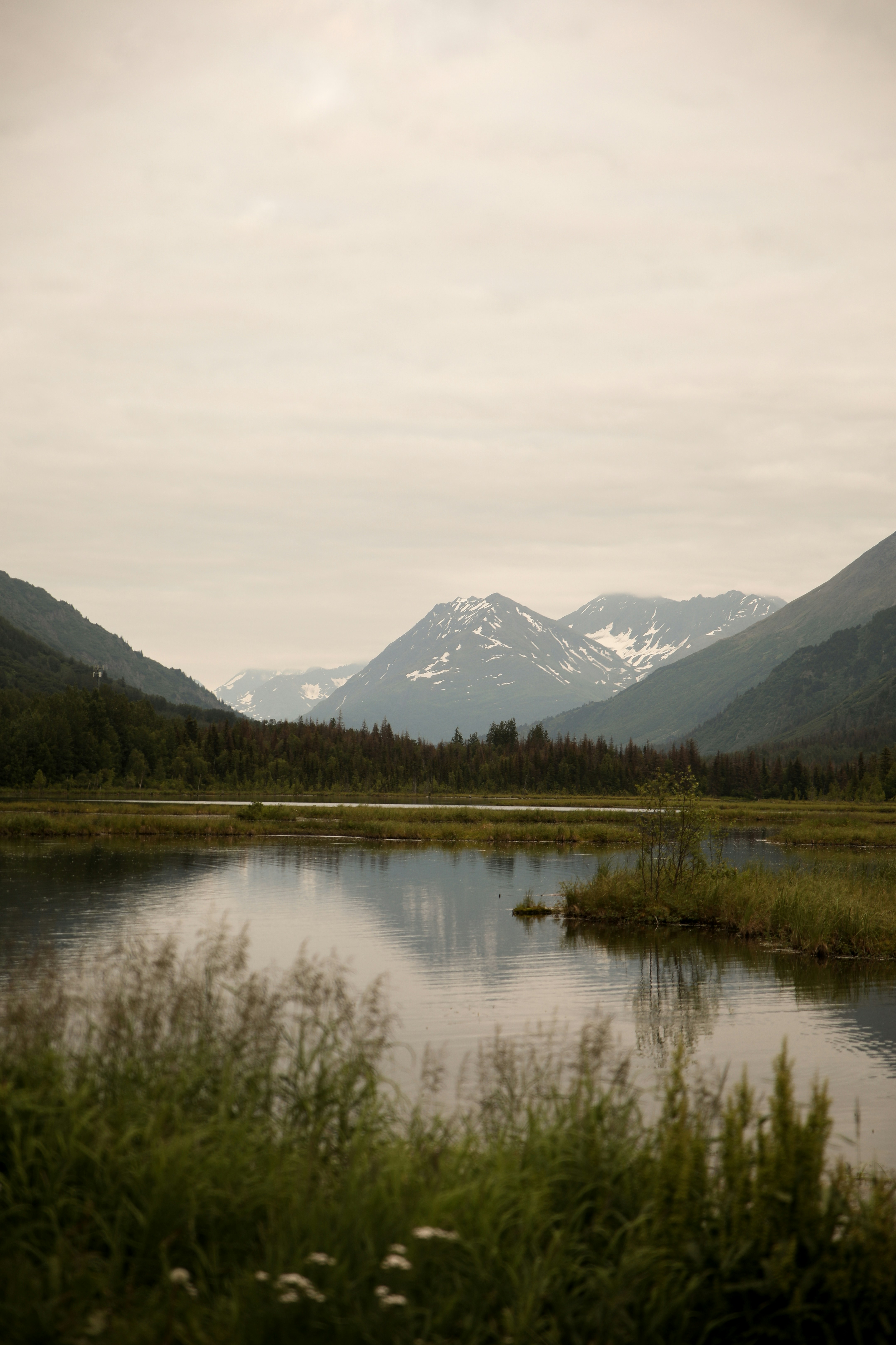 Calm lake reflects distant snow-capped mountains under cloudy sky.