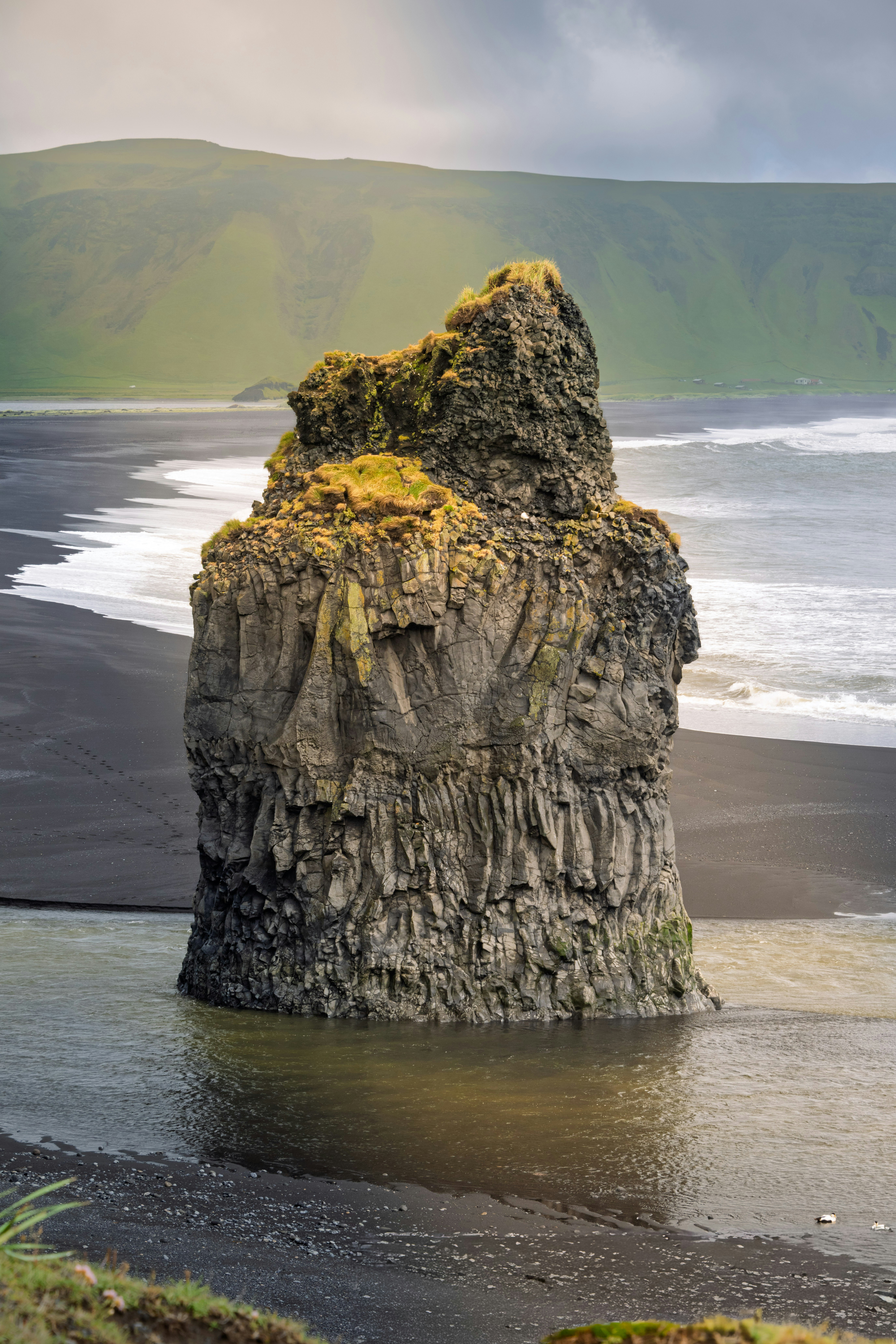 A lone basalt sea stack rises from the black-sand shore at Dyrhólaey, Iceland, with soft surf and mossy green cliffs under moody coastal light. | Tall rock formation on a black sand beach