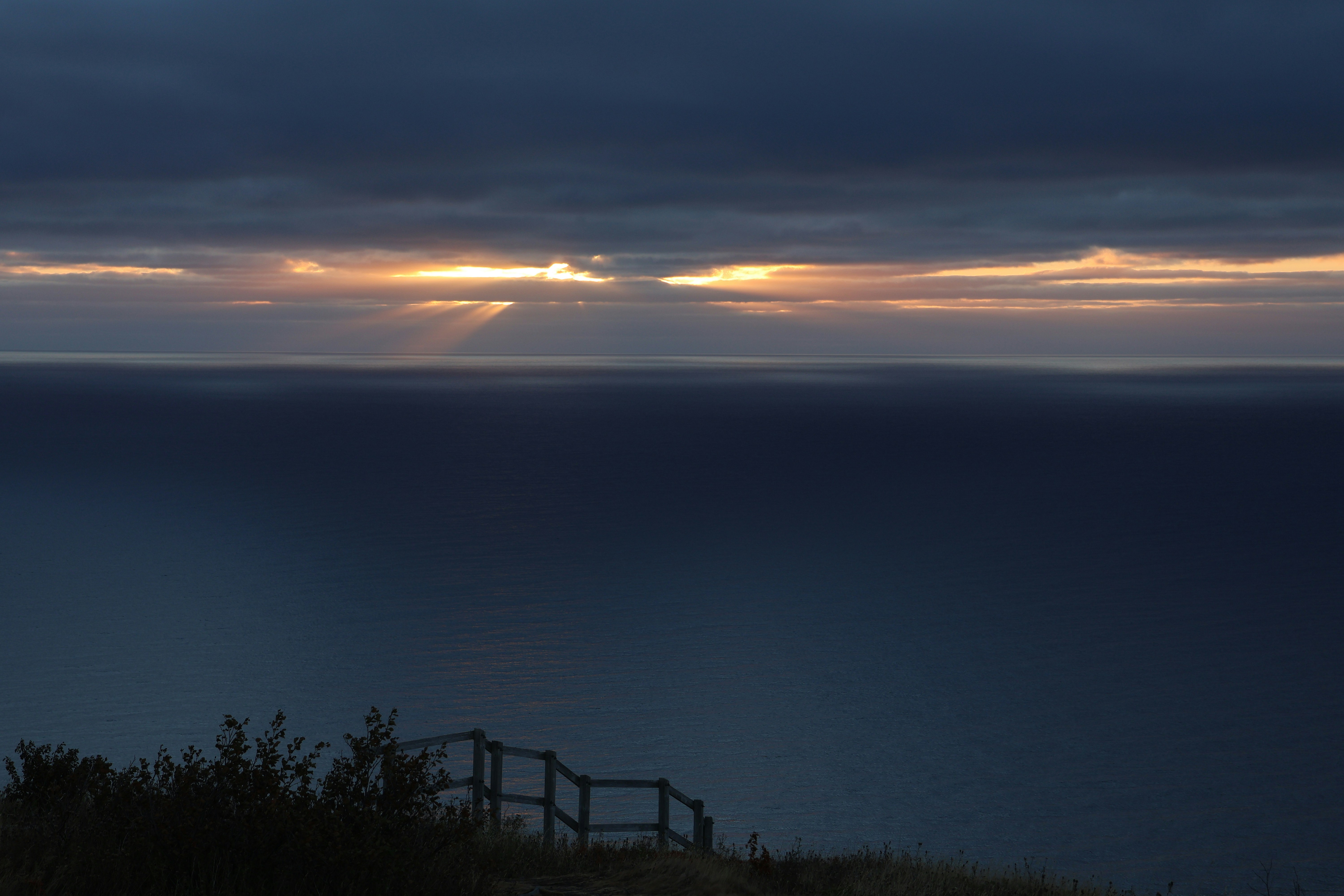 Soft rays of sunlight pierce through clouds, illuminating the tranquil ocean below. A wooden railing hints at a viewpoint, inviting contemplation.