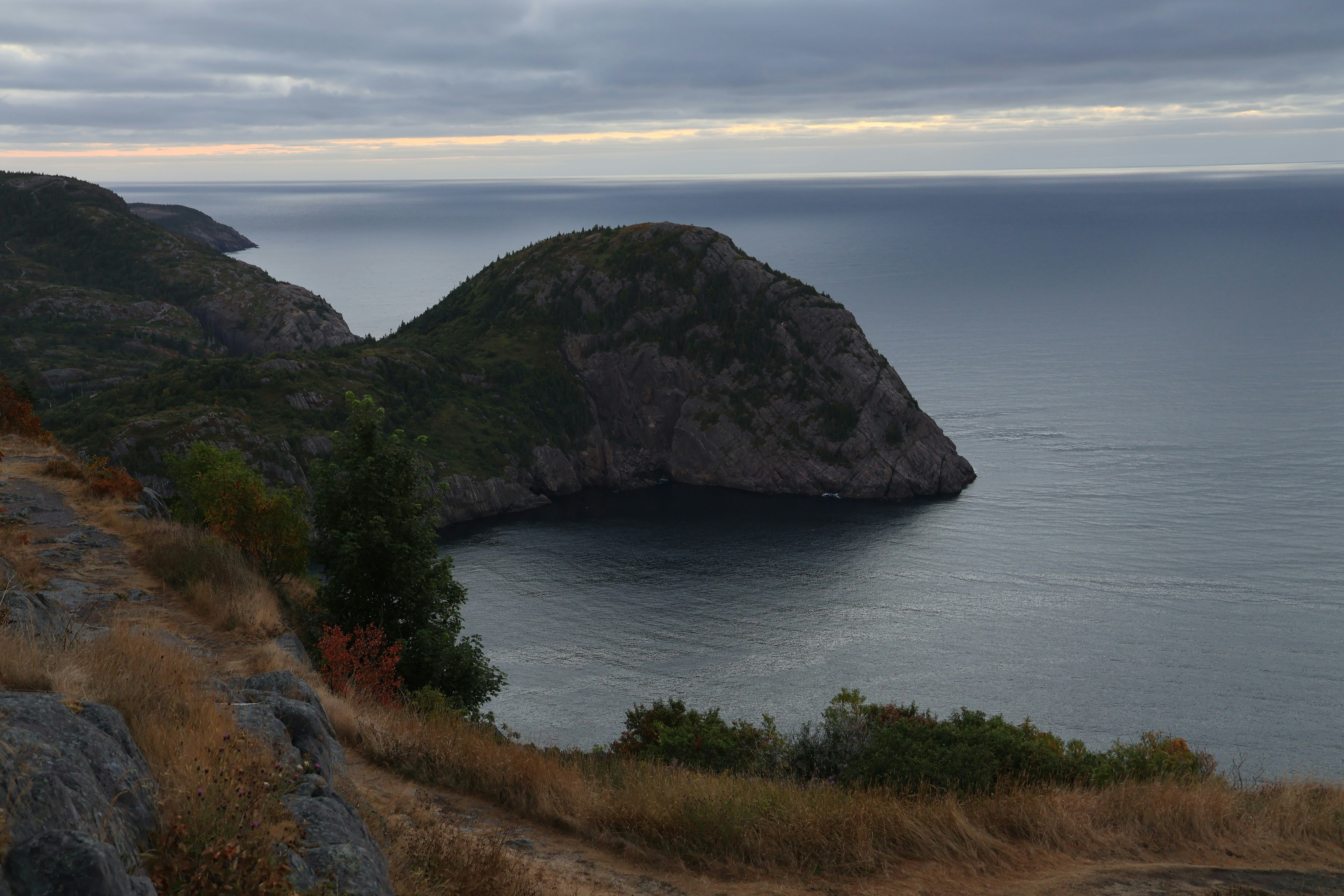 A serene coastal landscape featuring a rugged cliffside meeting the calm sea under a dramatic sky. The scene captures the tranquility of nature's contours.