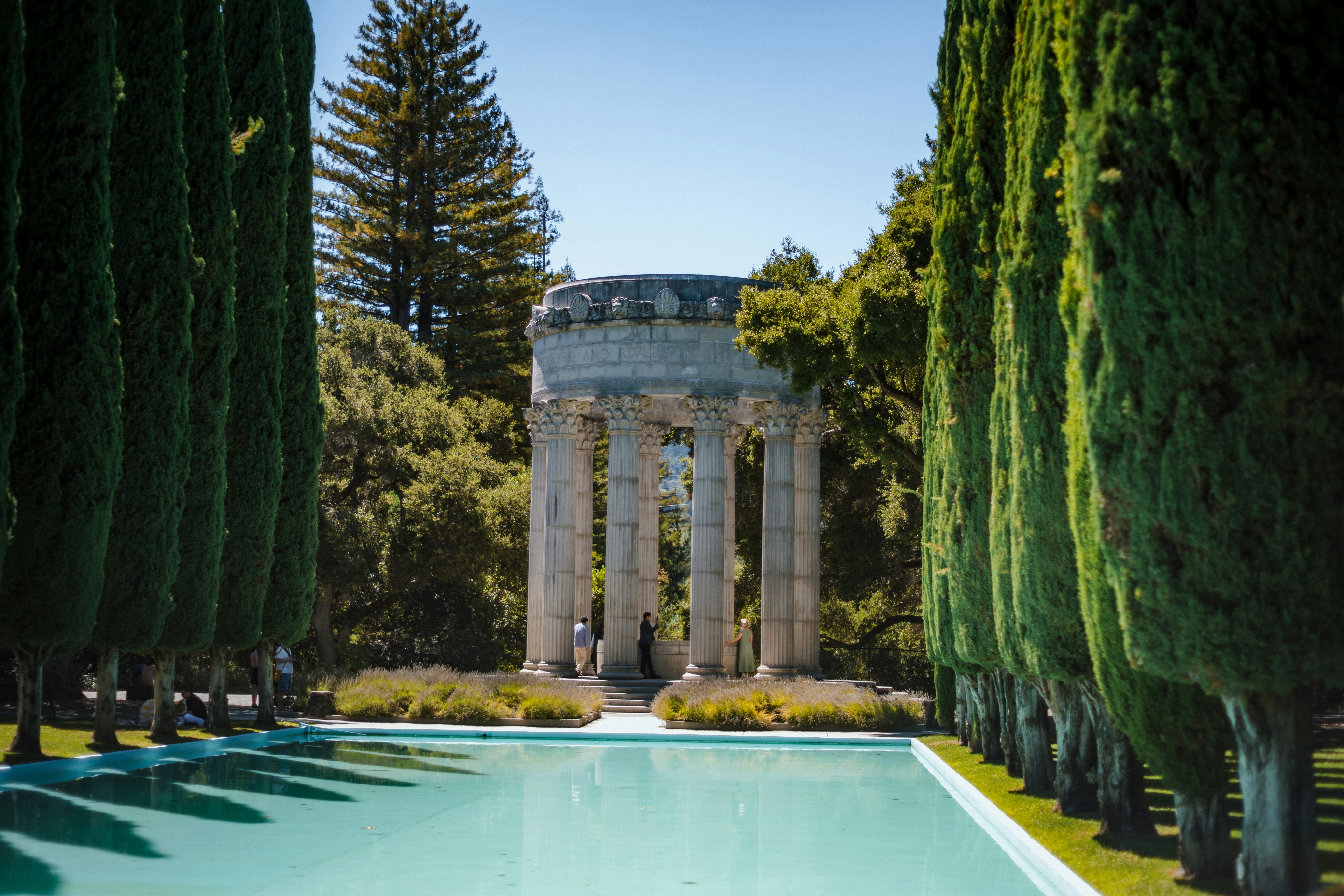 A serene garden scene featuring a classical stone structure framed by towering cypress trees, with a tranquil reflecting pool in the foreground.