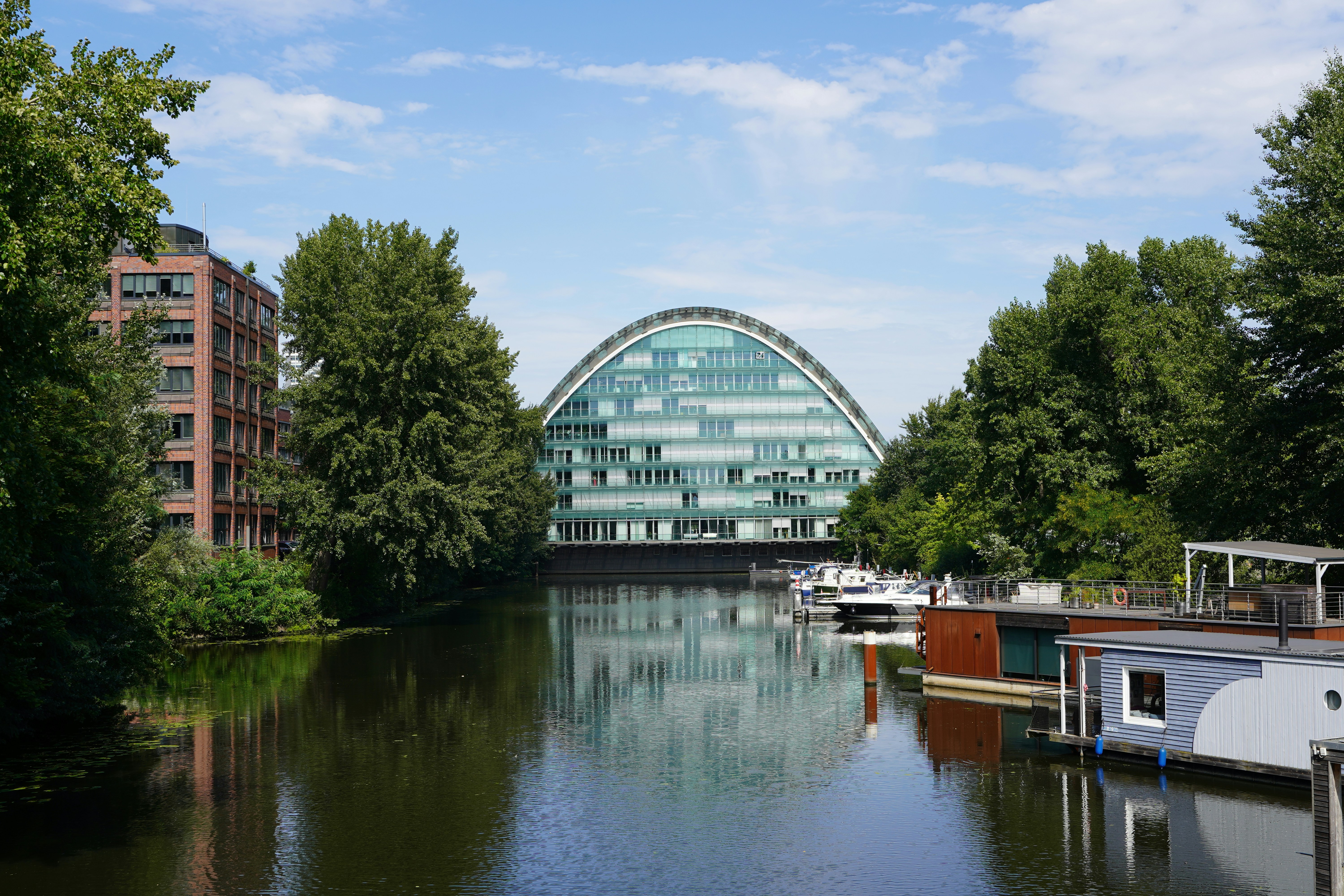 Contemporary building with a distinctive curved roof reflected in a tranquil waterway, surrounded by lush greenery.
