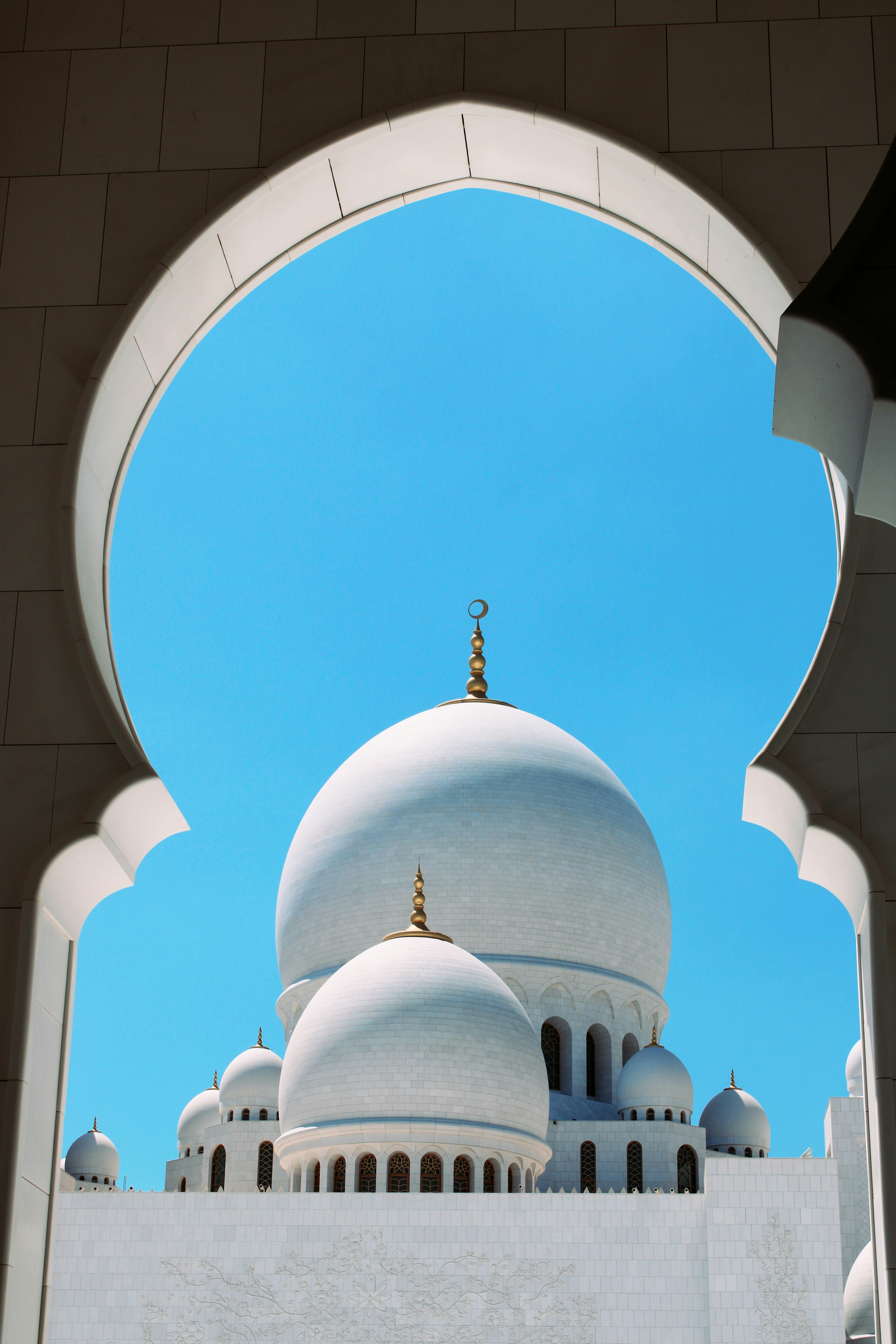 Elegant white domes framed by an ornate arch, set against a clear blue sky, showcasing intricate Islamic architecture.