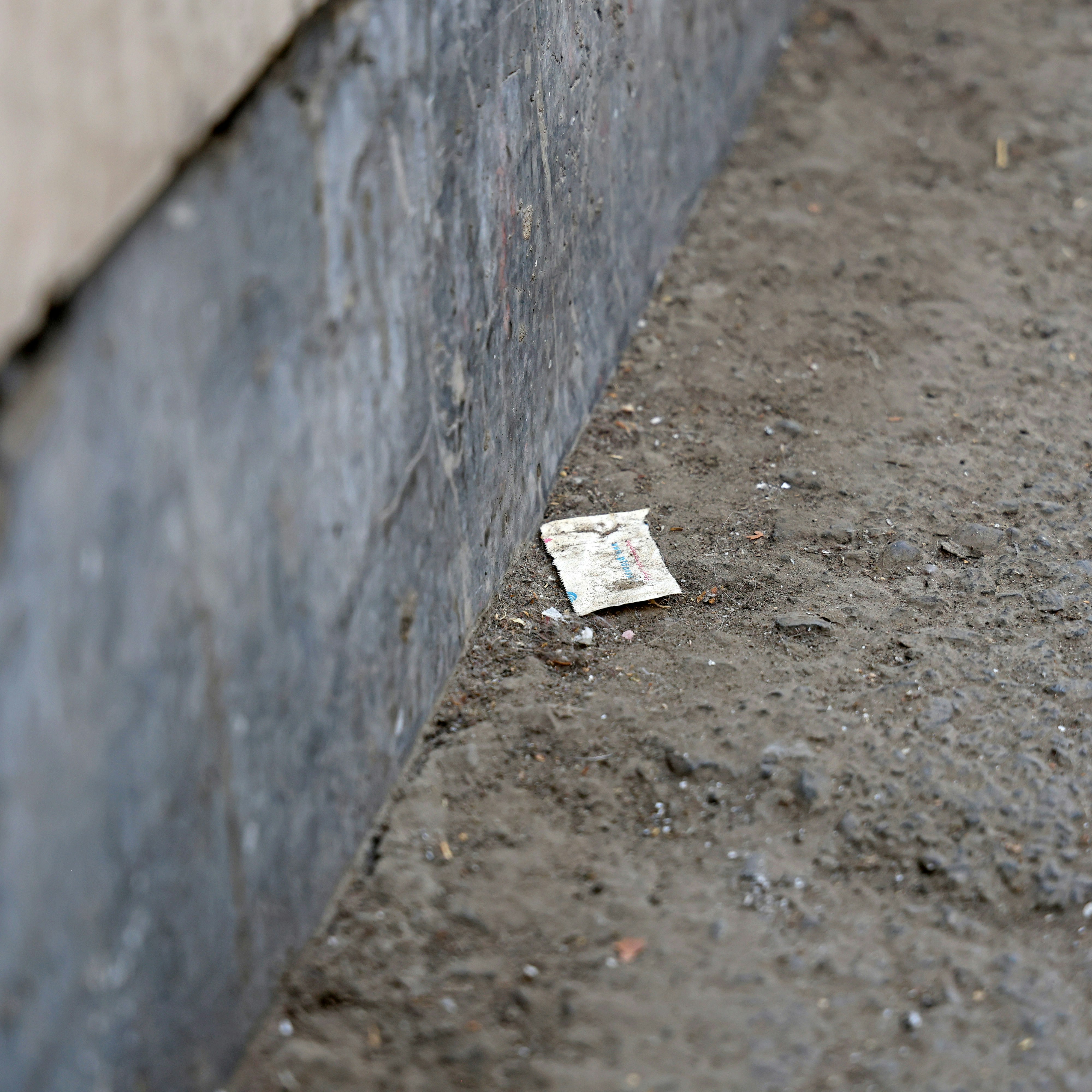A small piece of paper, a purchase receipt, lying on the corner of the sidewalk next to a wall in an alley. | Small wrapper on dirty ground next to wall