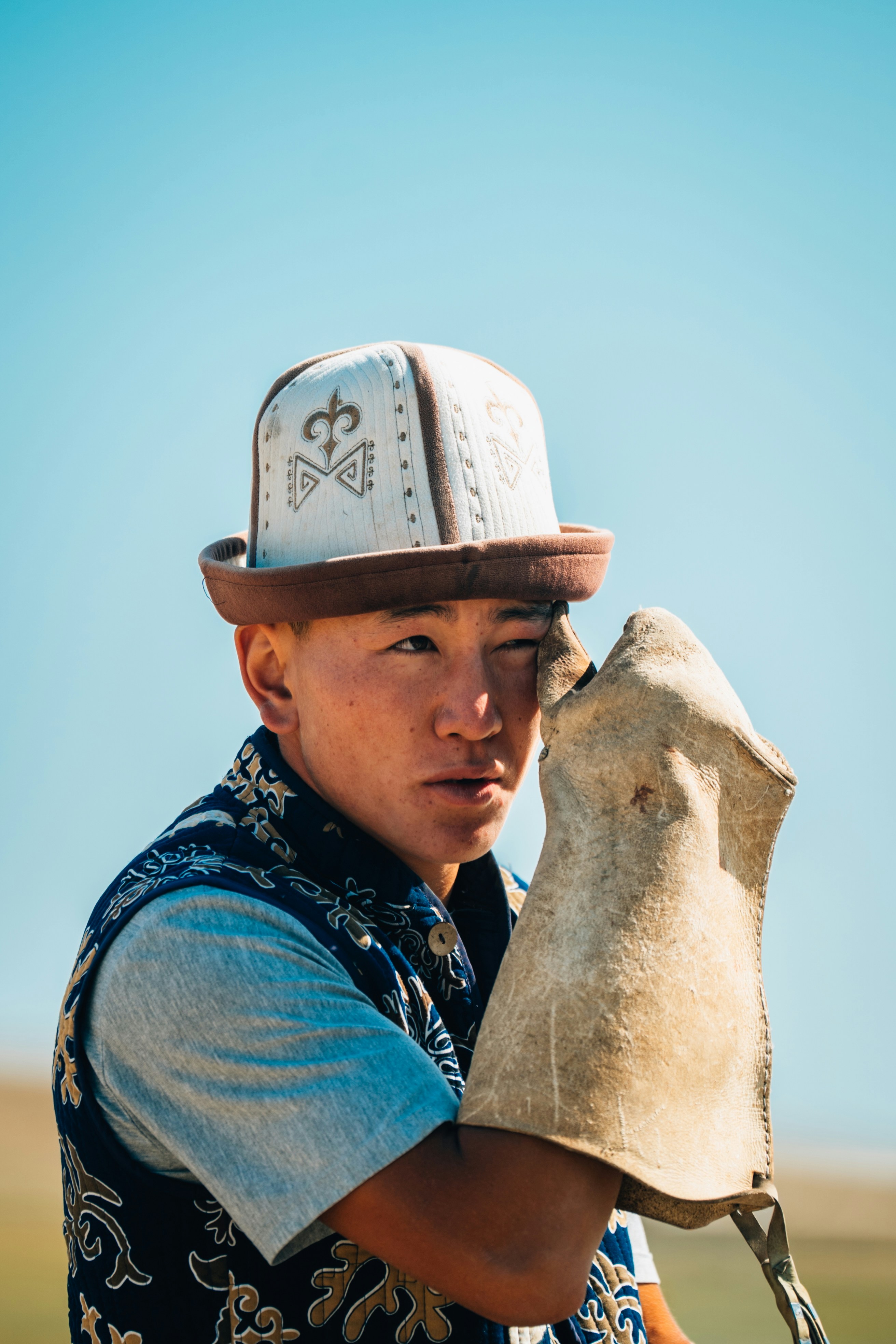 Young falconer preparing to train his bird, wearing traditional attire and a protective glove. The clear blue sky serves as a backdrop.