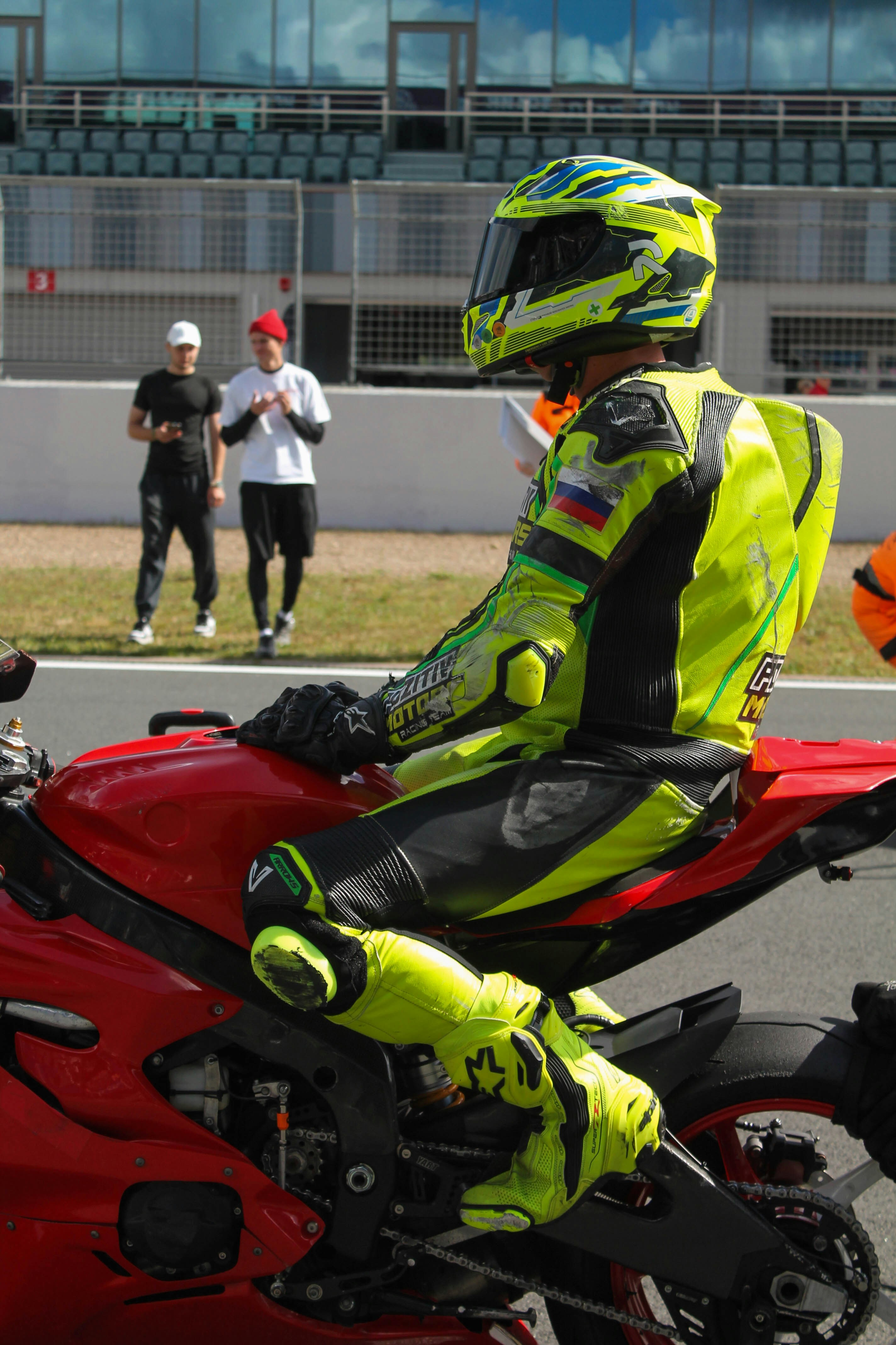 Motorcycle racer in vibrant gear poised on a red bike, surrounded by spectators at a racetrack.
