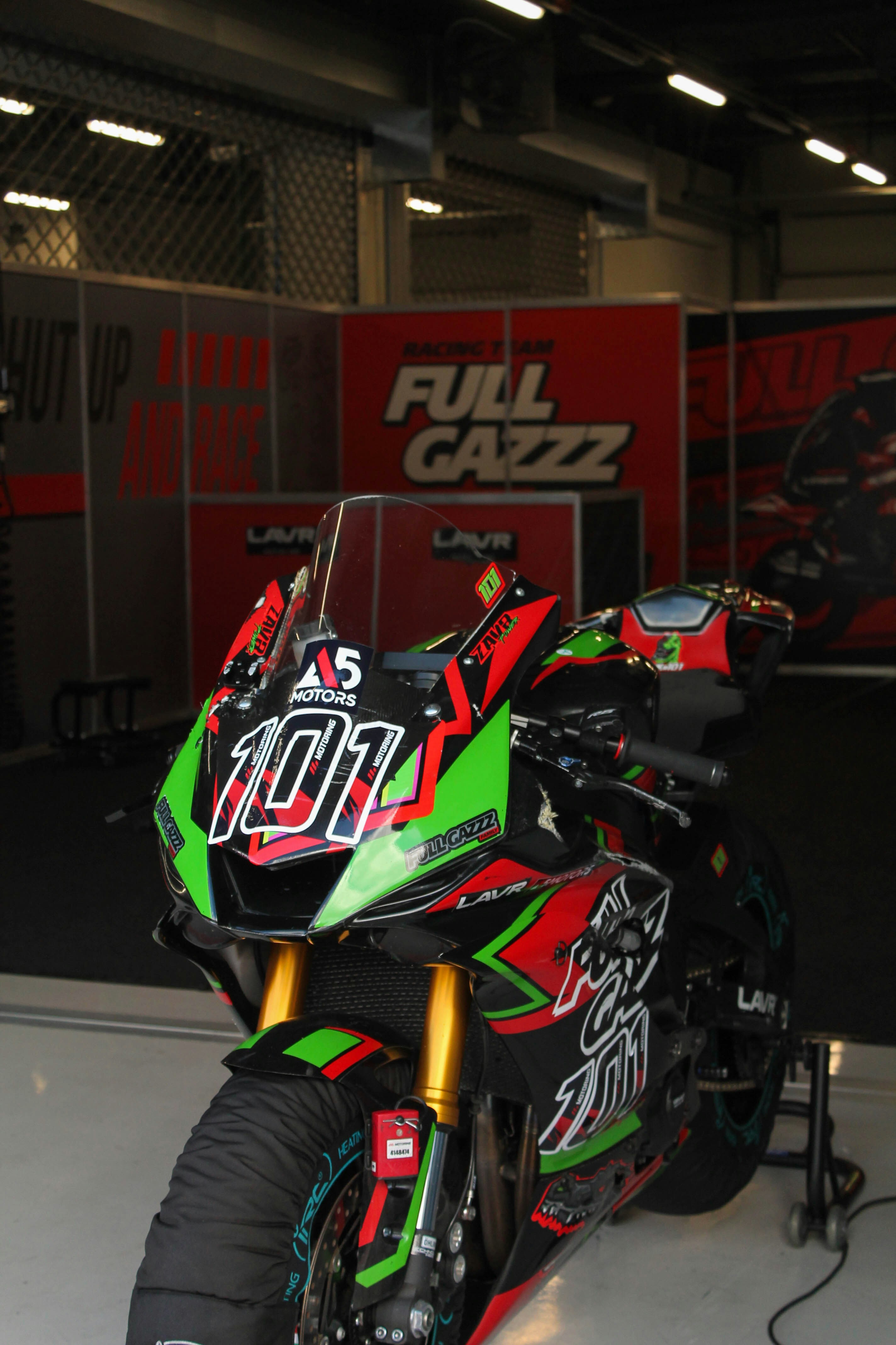 A brightly colored racing motorcycle adorned with bold graphics and the number 101, positioned in a garage setting with promotional banners in the background.