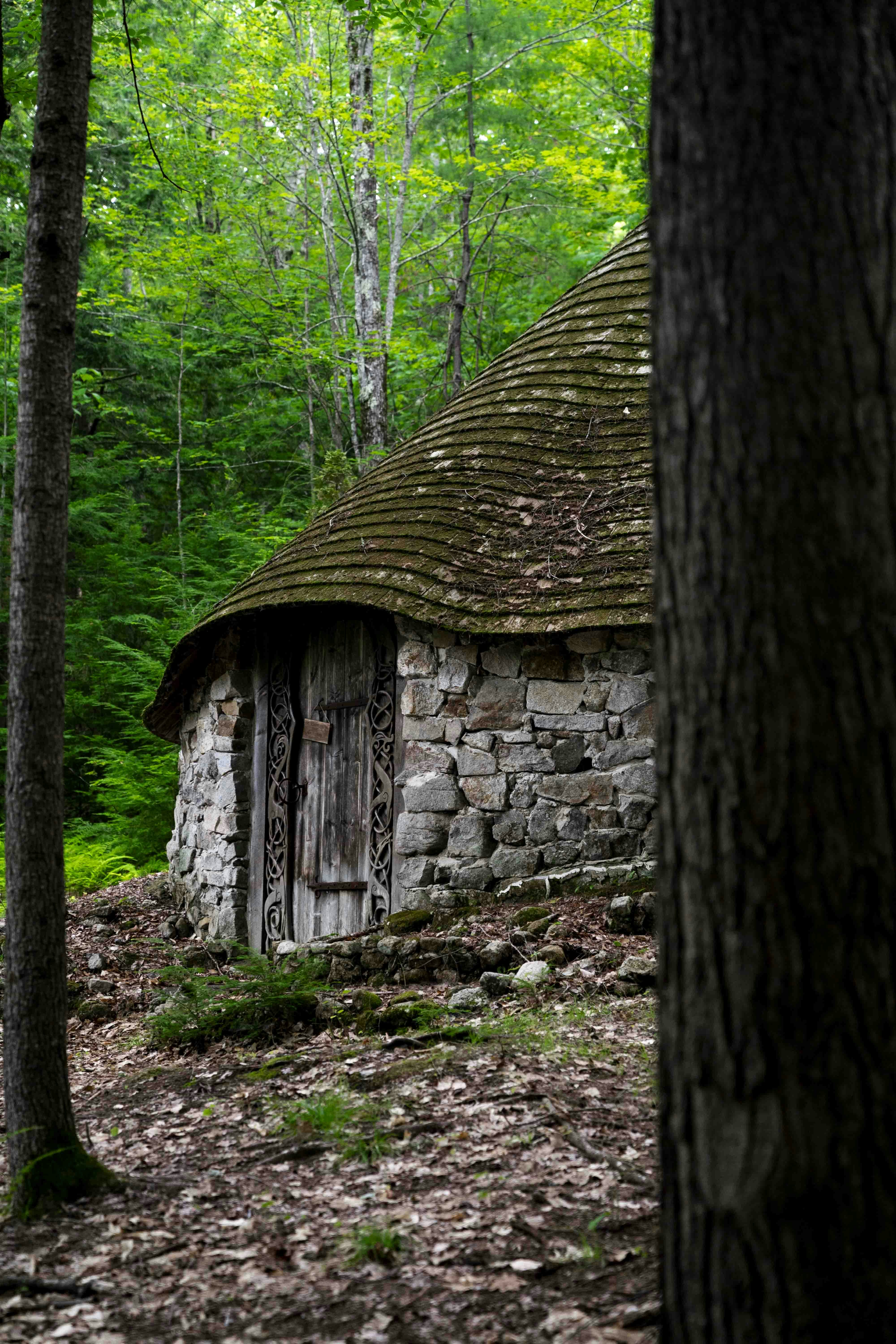 A rustic stone structure nestled among lush greenery, featuring a distinctively crafted wooden door and a conical roof. Nature envelops the scene, hinting at stories untold.