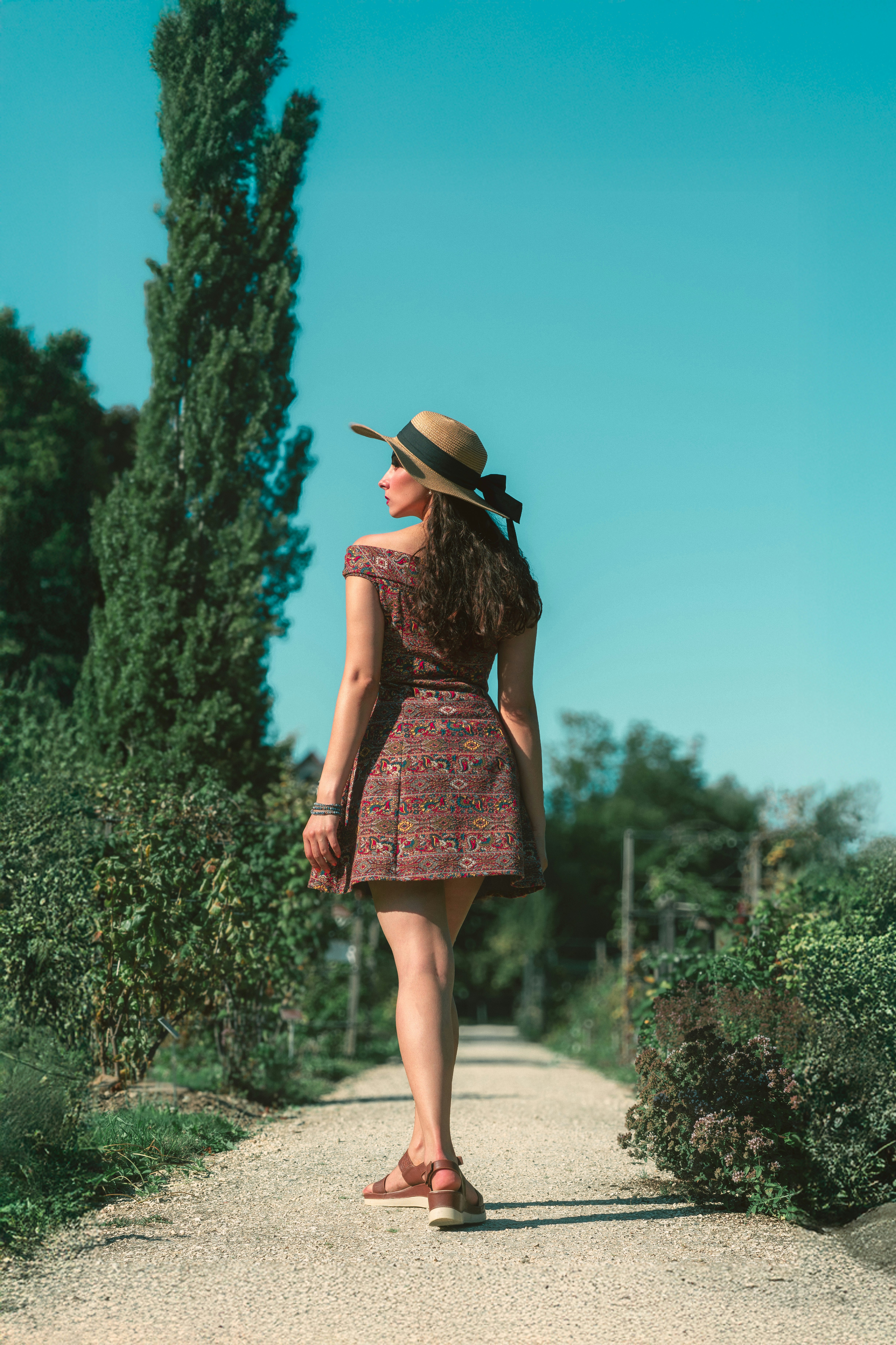 Woman in a patterned dress walking along a sunlit path flanked by greenery and trees.