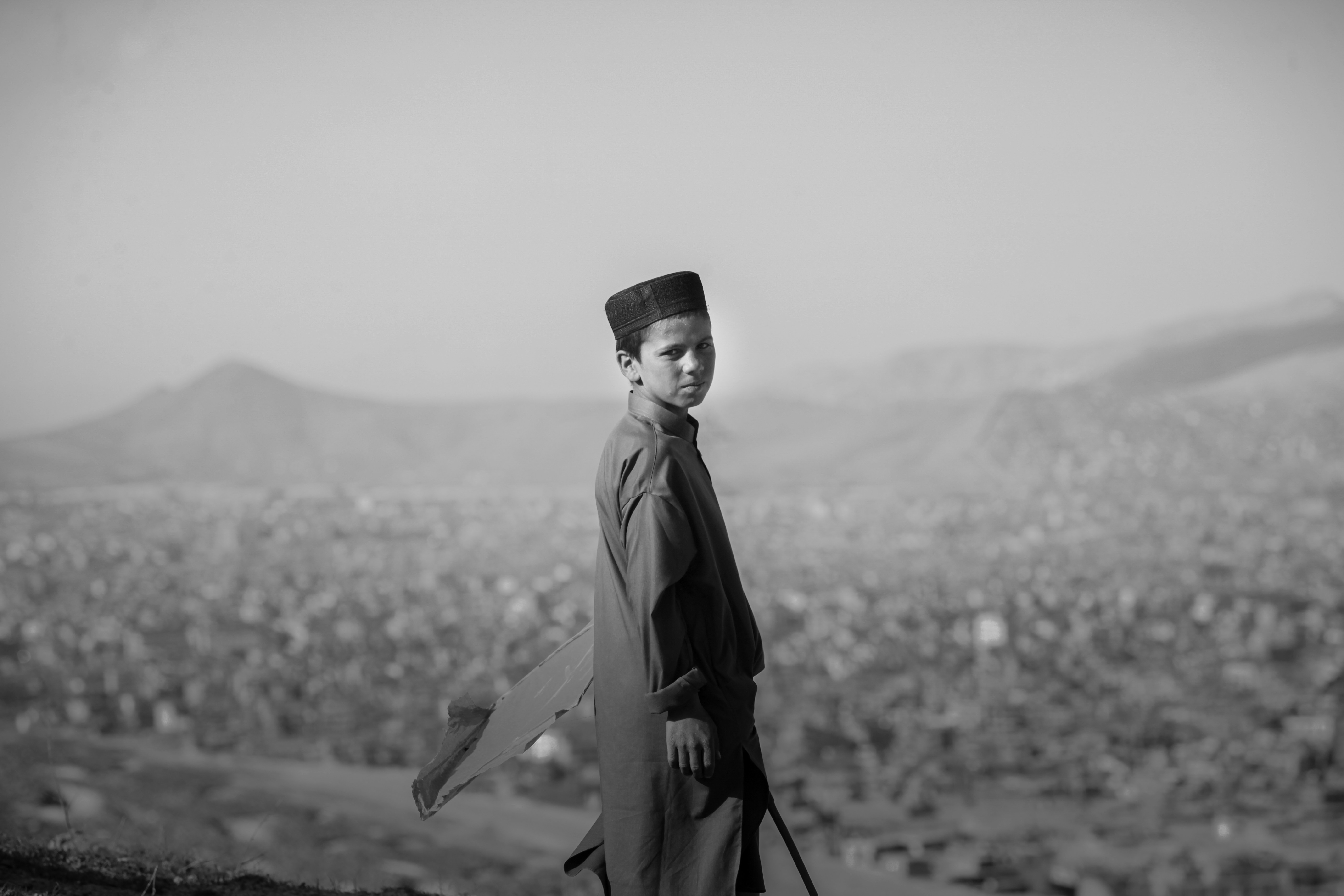 Photo of an Afghan boy grazing his sheep in the hills of Kabul. | Young boy in traditional clothing looks over cityscape