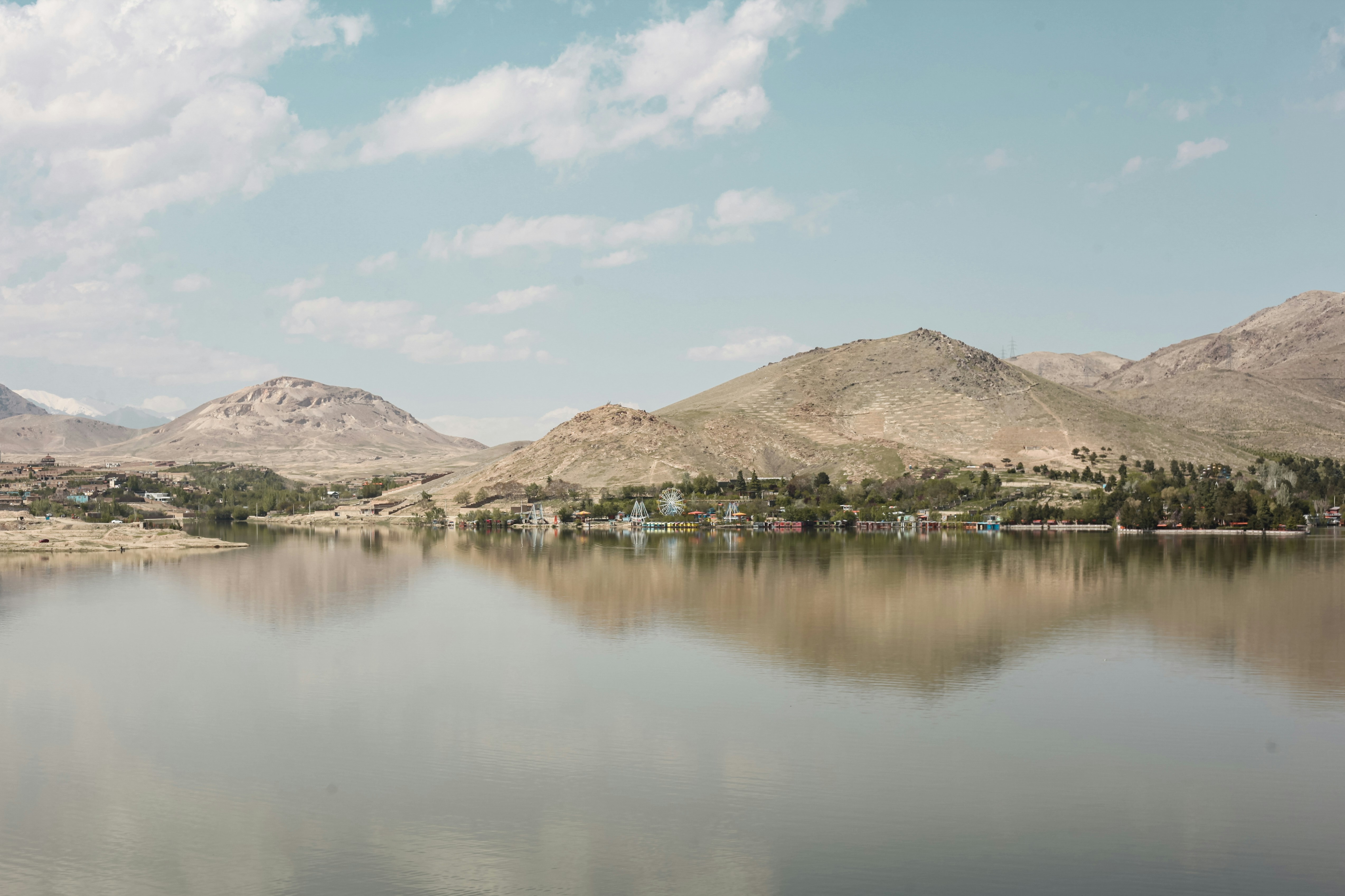 Qargha Reservoir, also known as Qargha Lake, is a popular recreational area near Kabul, Afghanistan. It's a man-made lake formed by the Qargha Dam on the Paghman River. Initially built for irrigation in the 1930s, it evolved into a recreational hub with activities like boating, fishing, and picnicking. | Calm lake reflecting distant mountains and buildings