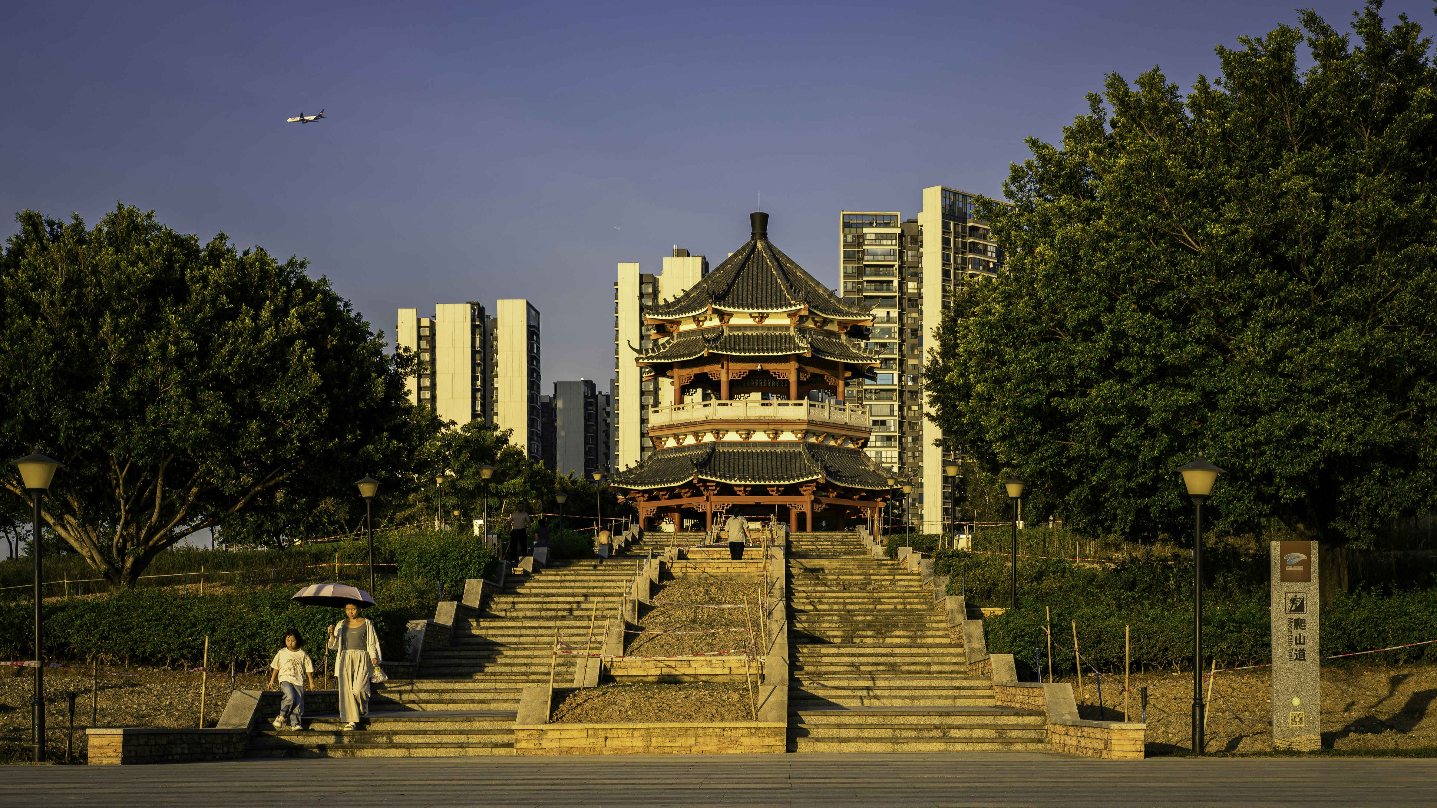 A traditional Chinese pagoda stands majestically at the top of a grand stone staircase, bathed in the warm, golden light of the late afternoon sun. This serene scene, set within a lush park, presents a striking juxtaposition of old and new, as modern high-rise buildings tower in the background against a clear blue sky. People, including a mother and child with a parasol, can be seen enjoying the tranquil atmosphere, highlighting the harmony between cultural heritage and contemporary urban living. | Traditional pavilion on stone steps with modern buildings behind