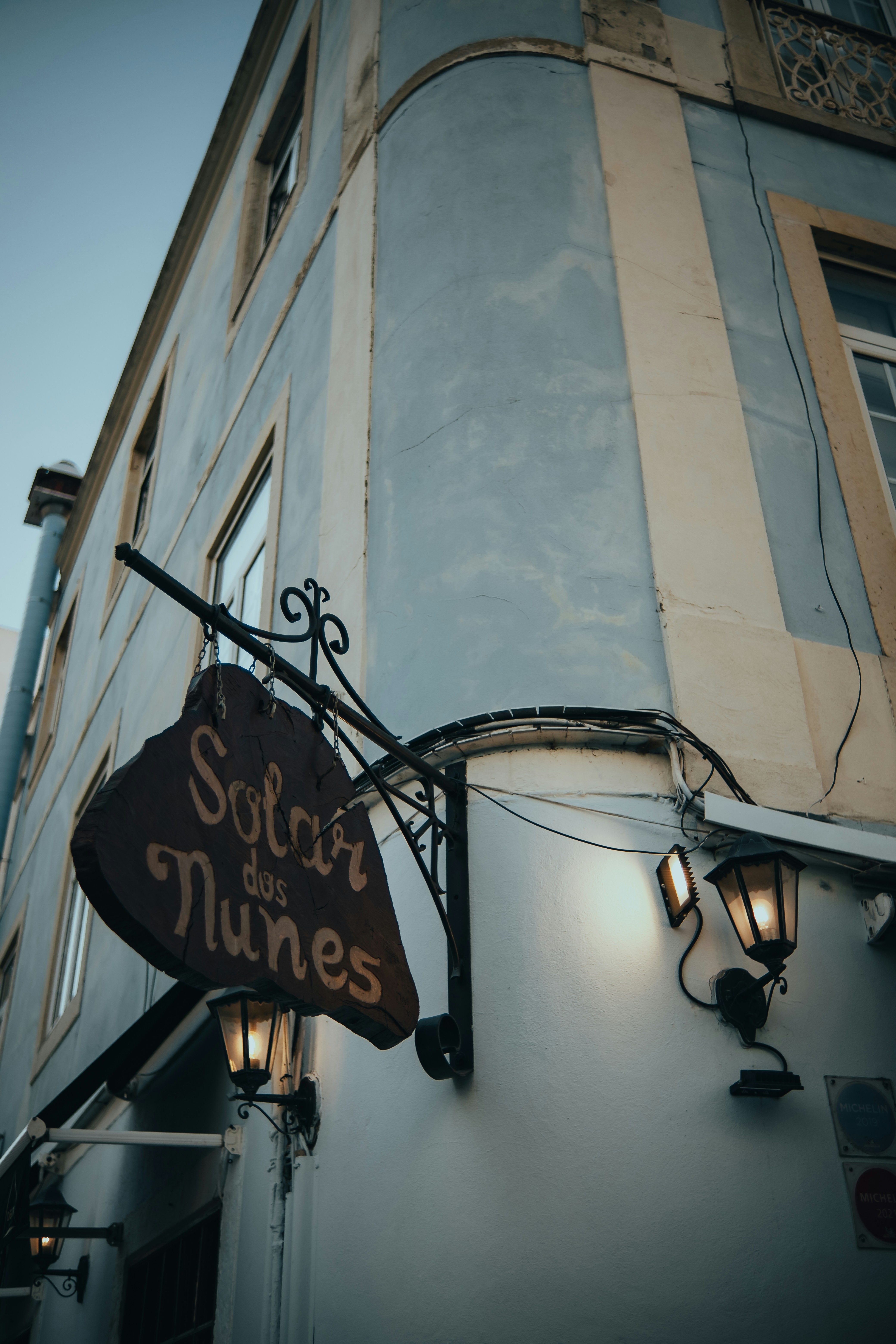 Vintage wooden sign hanging from a wrought iron bracket, nestled against a pastel blue wall, illuminated by warm street lamps.