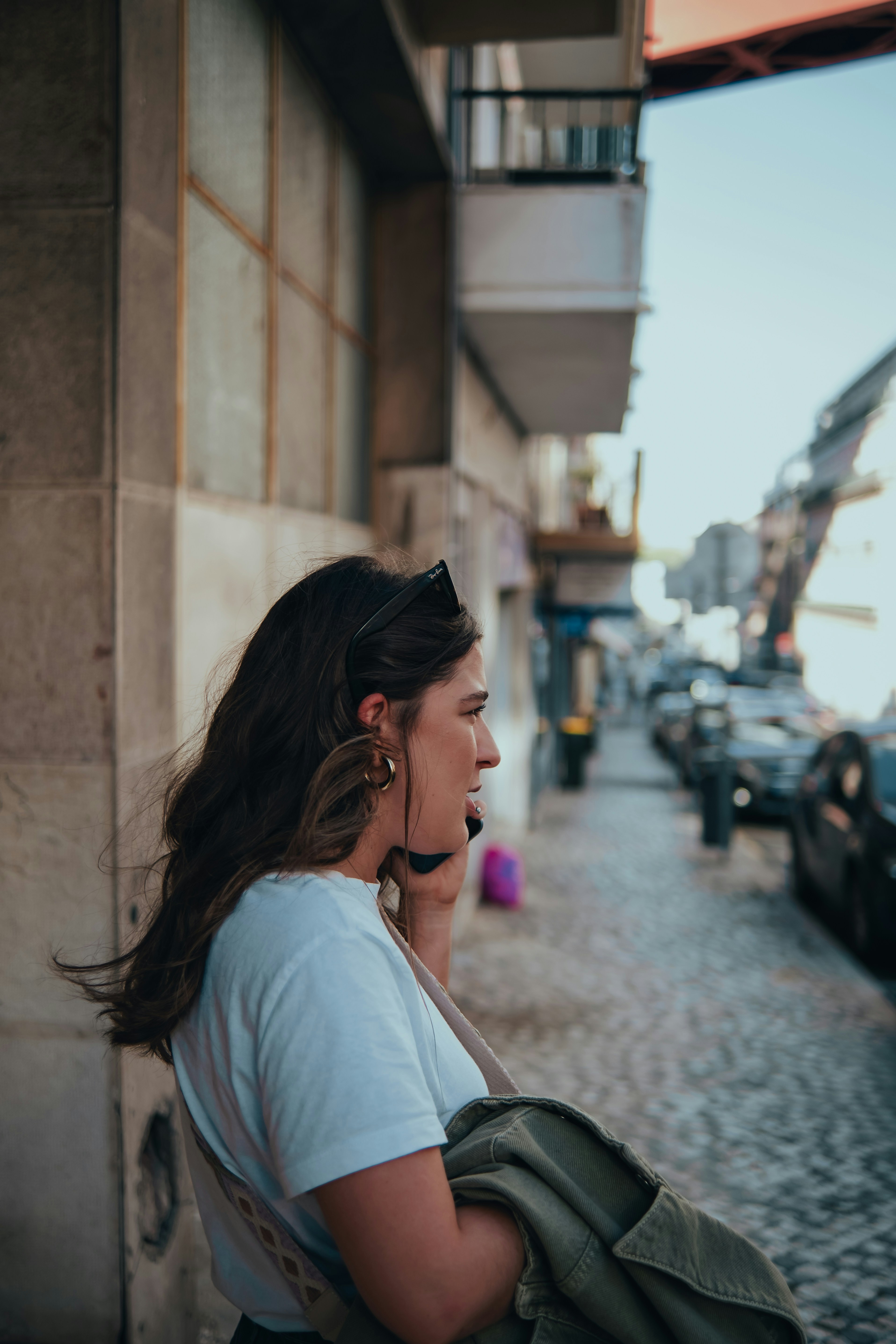 Young woman engaged in a phone call while walking along a cobblestone street, framed by urban architecture and parked cars.